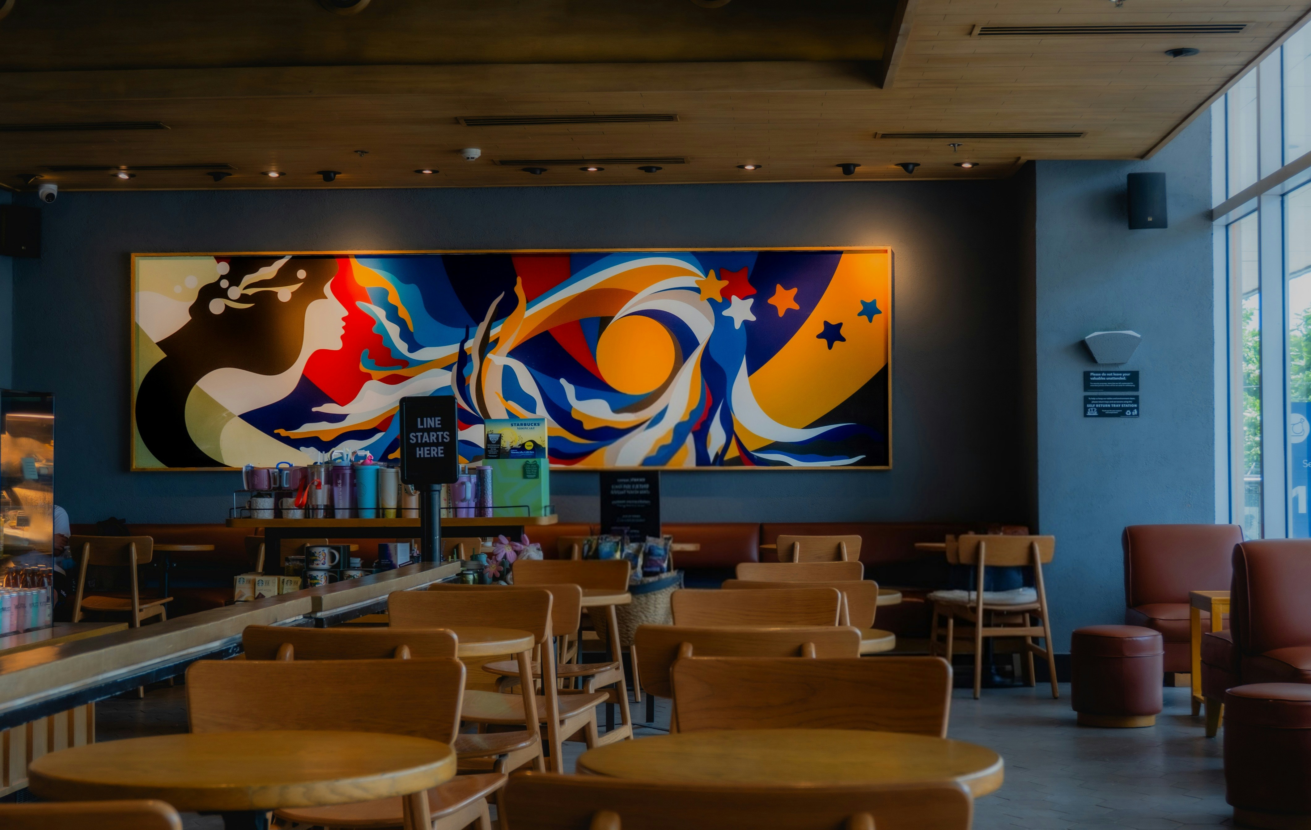 Warm, empty cafe with wooden tables and chairs set before a bold mural, hinting at a lively outdoor scene.