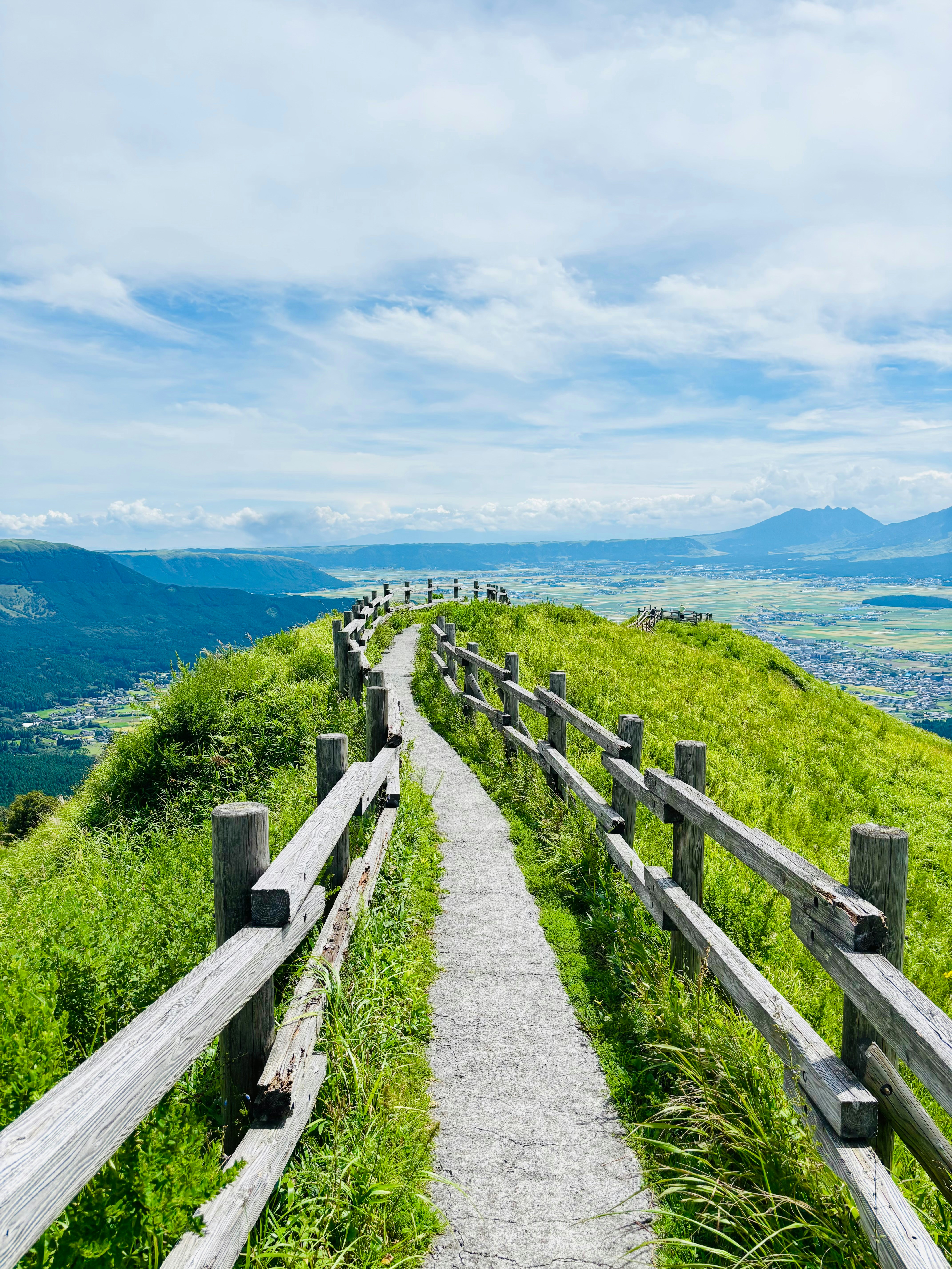 A breathtaking panoramic view of the lush green valleys and majestic mountains of Aso, taken from the scenic pathway at Nishiyunouraenchi Observatory under a beautiful blue sky.