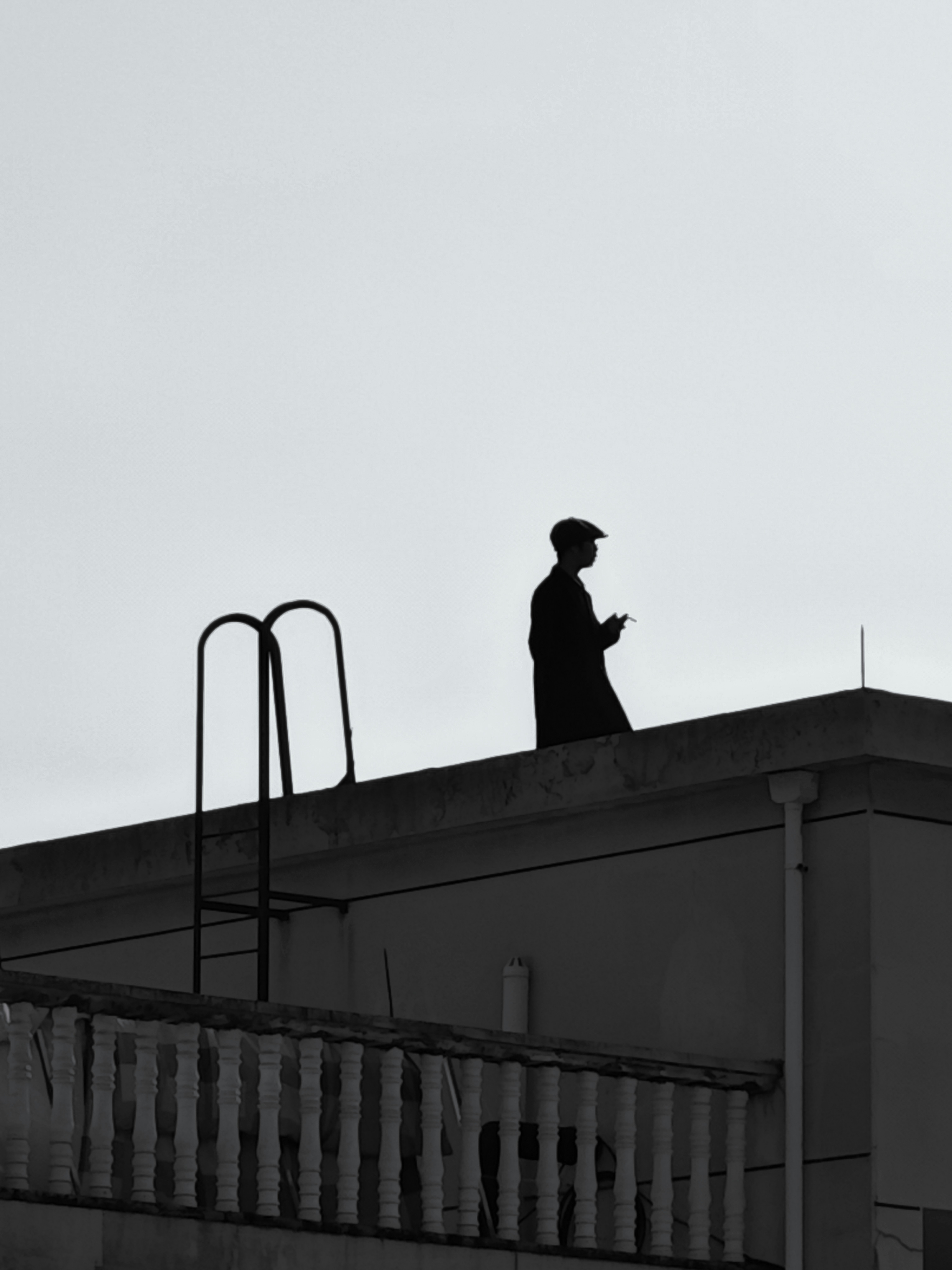 A lone figure stands on a rooftop against a pale sky, silhouette framed by architectural elements and a ladder railing, conveying quiet urban solitude. This is a monochrome photograph.