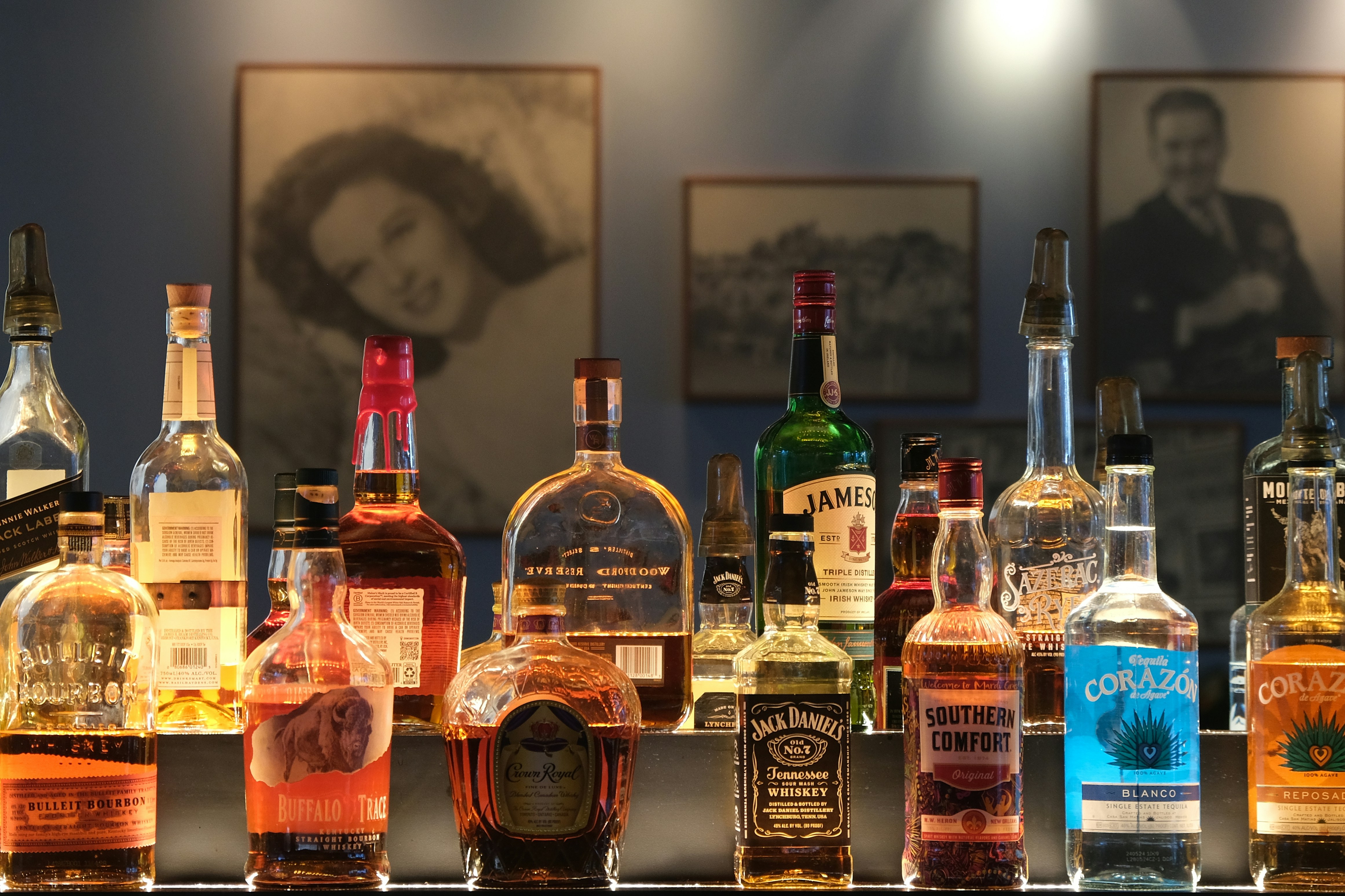 Row of assorted liquor bottles on a backlit bar, with warm amber tones and varied label designs.