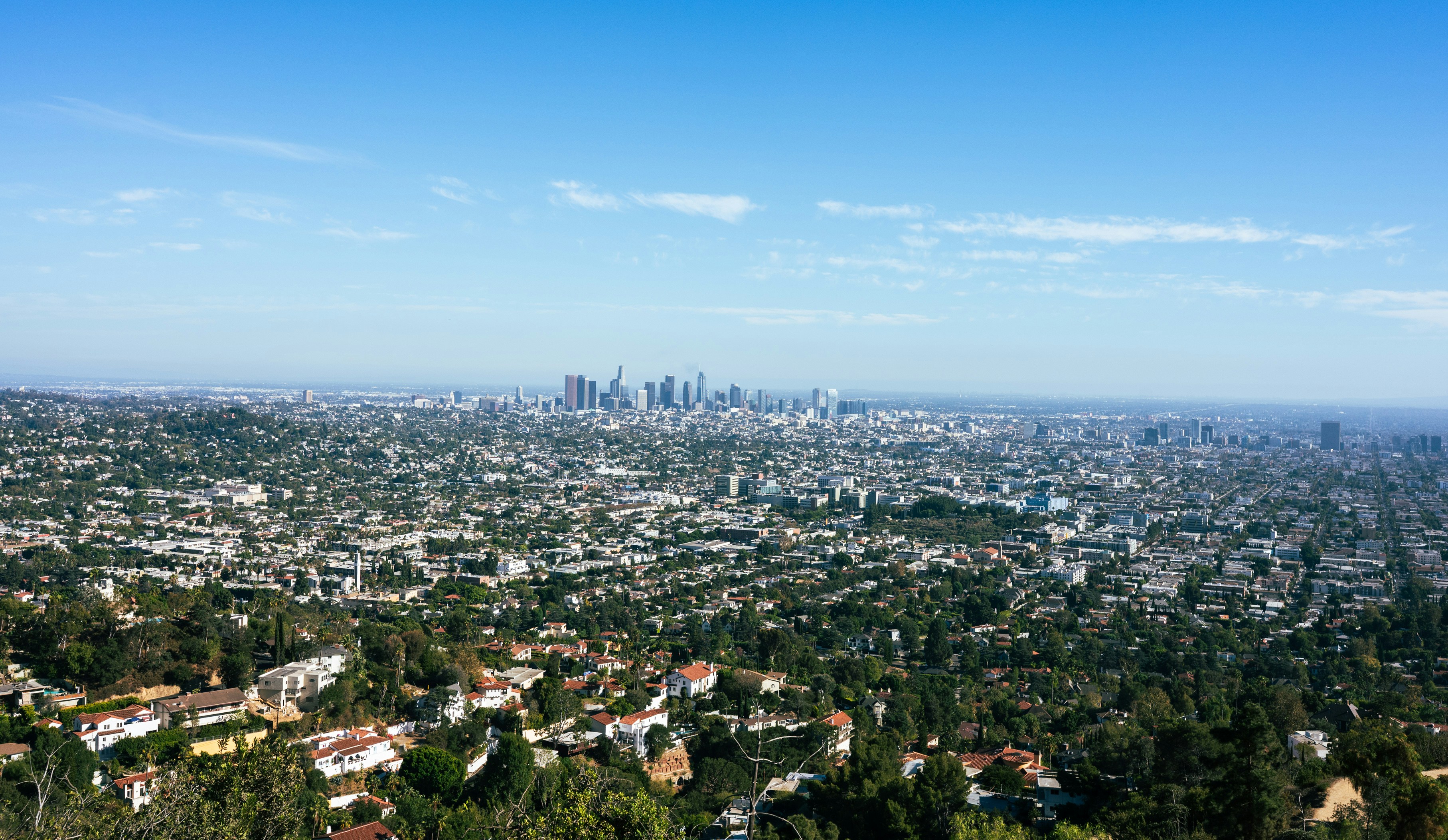 A sweeping aerial view of a sprawling city spreading from green hills to a distant skyline, with a clear blue sky above. The shot emphasizes the expansive urban landscape and distant downtown silhouette.
