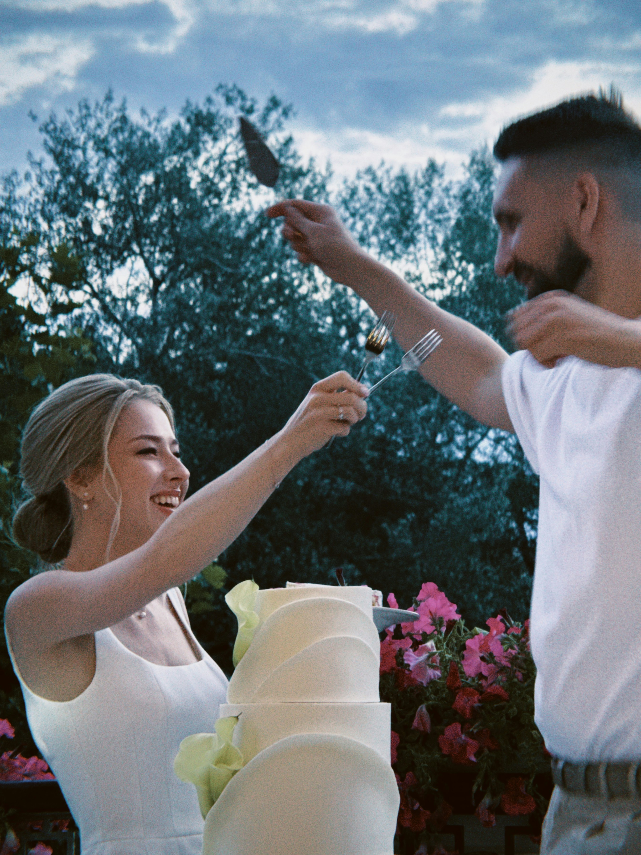 A couple toasting with forks and a cake on an outdoor terrace, with greenery and flowers in the background.