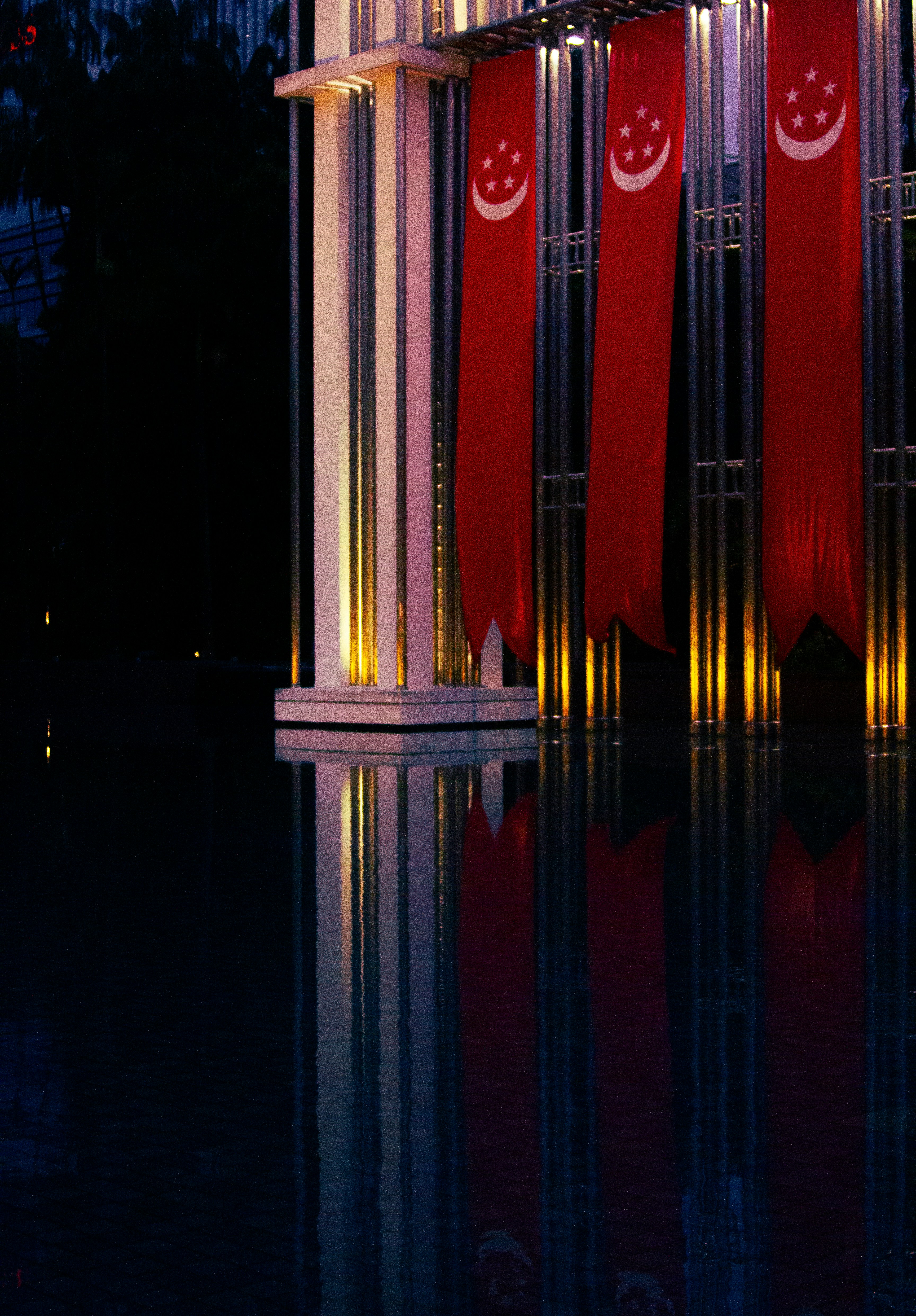 A dramatic night view of the illuminated Festival Arch at Istana Park, Singapore, featuring vibrant red national banners with the crescent moon and stars, beautifully reflected in the still surface of the park's water feature.