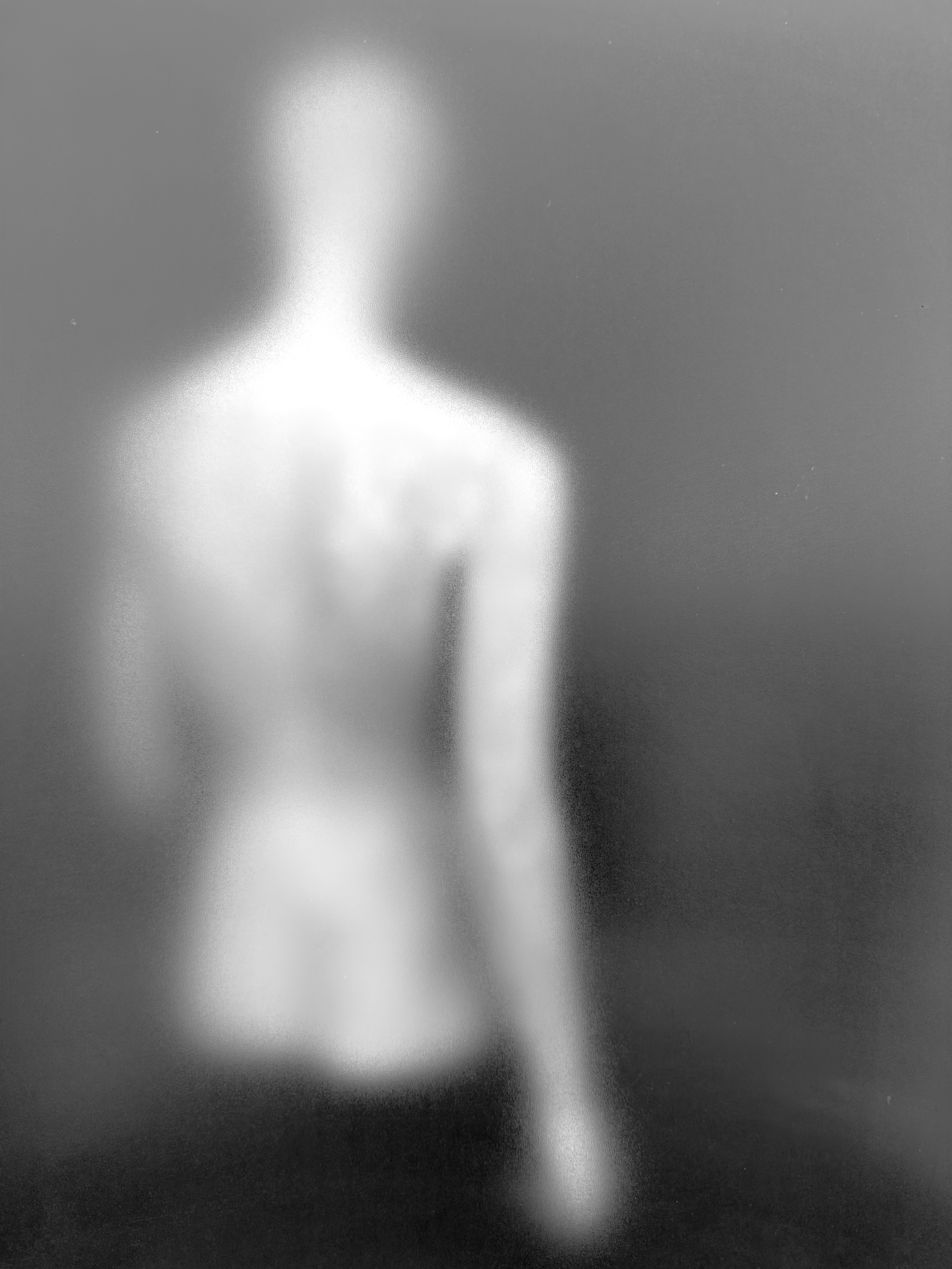 A blurred, monochrome silhouette of a standing human figure seen through a translucent surface, emphasizing form over detail. The image reads as an abstract study of presence and indistinct identity.