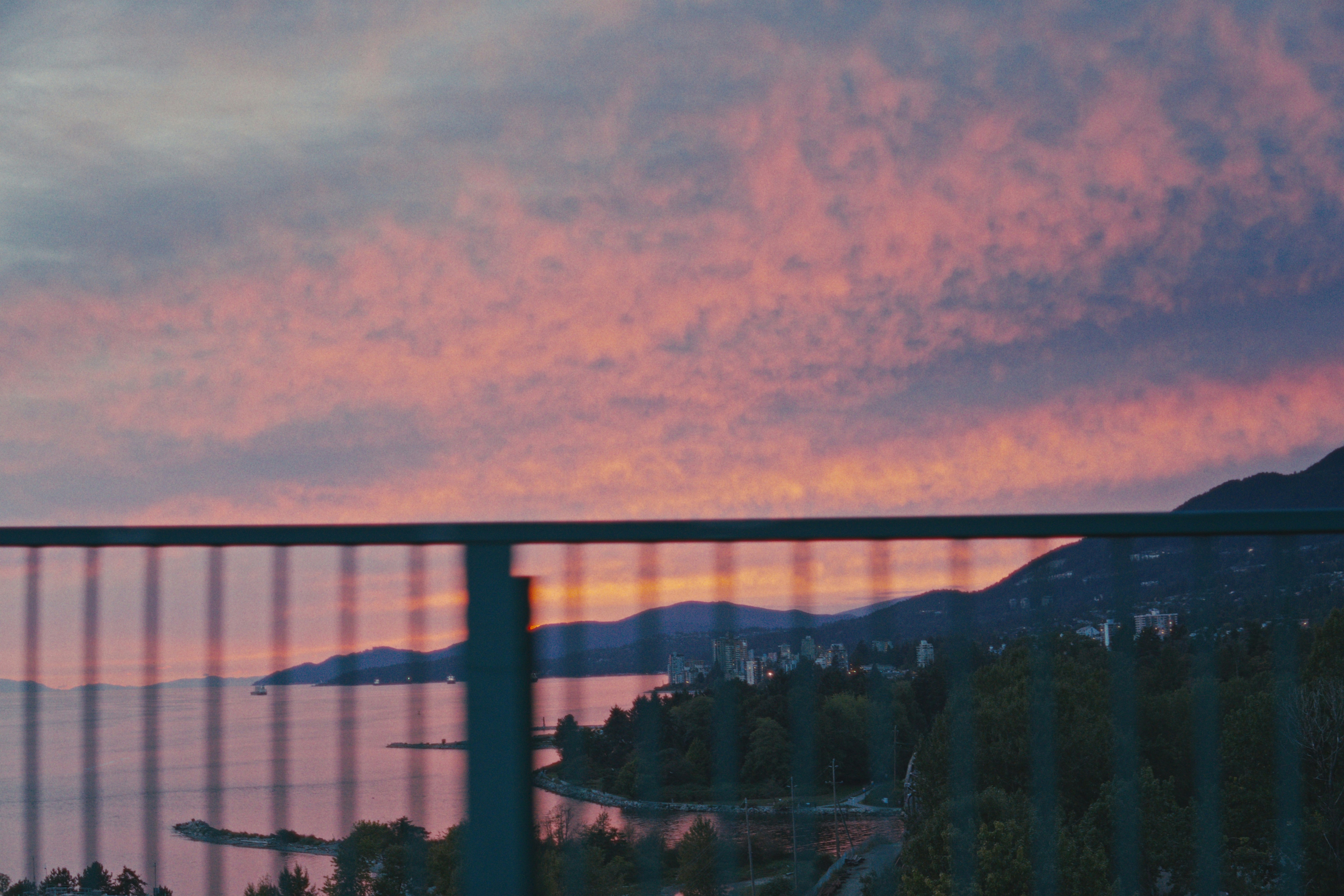 Soft sunset hues over distant hills viewed beyond a railing, with the silhouette of coastline and distant vegetation. A coastal overlook scene captured with a shallow-depth feel.