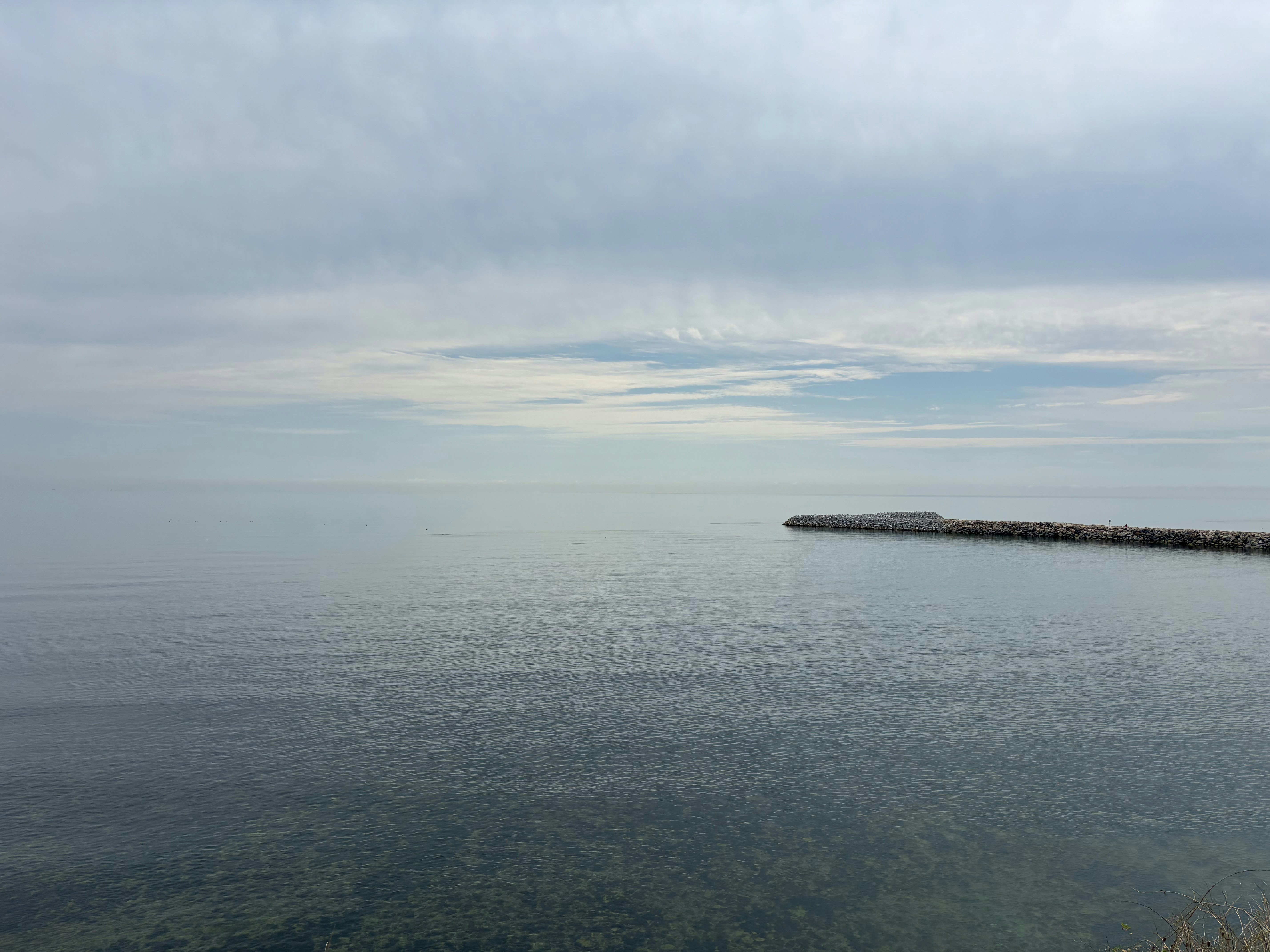 Calm seascape showing a tranquil shoreline with a distant breakwater stretching into the horizon under soft, overcast skies. A clear gradient of water depth meets the sky, emphasizing serenity and vastness.