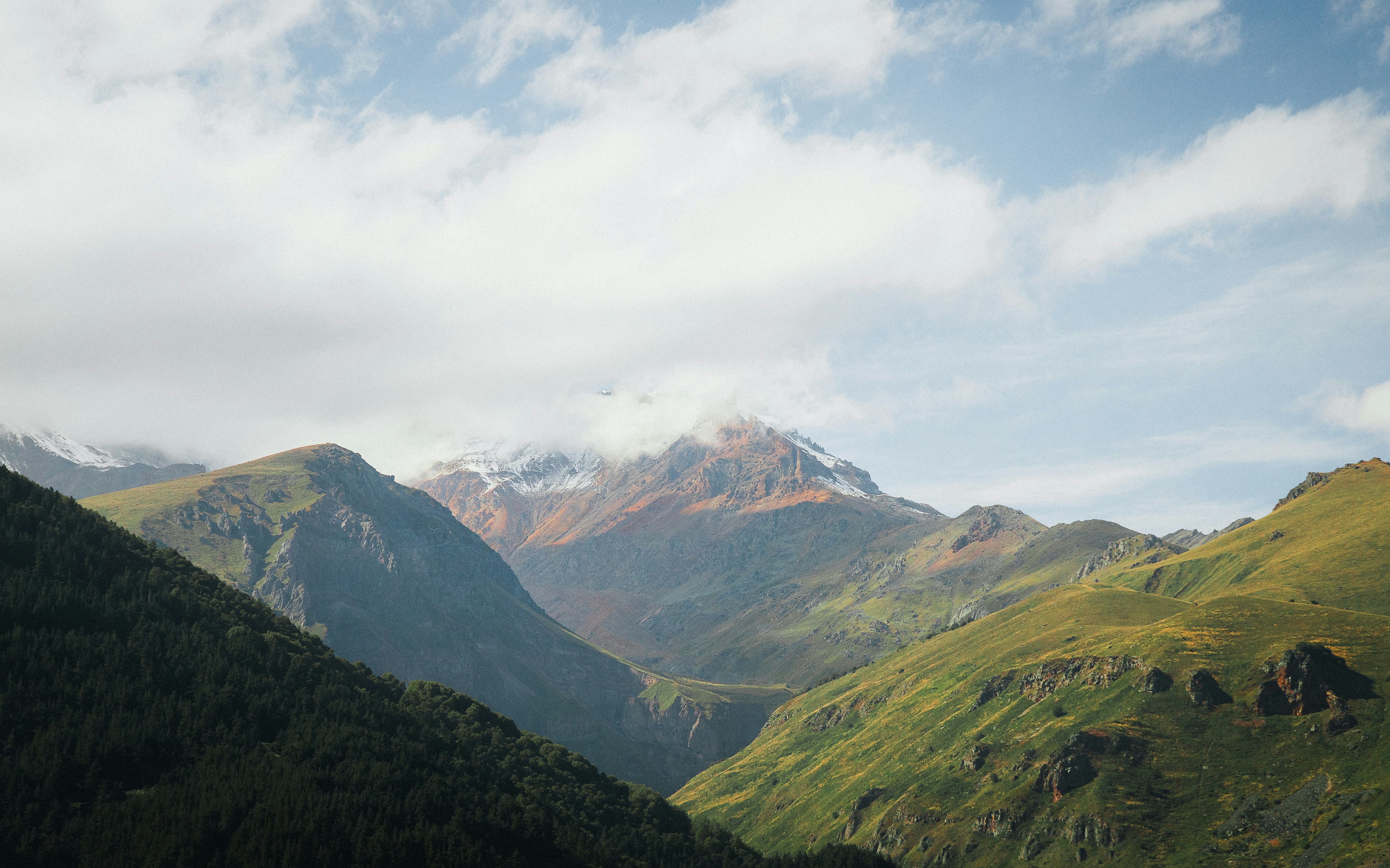 Rolling green valleys and rugged mountains under a blue sky with scattered clouds; a panoramic alpine landscape emphasizing depth and scale.