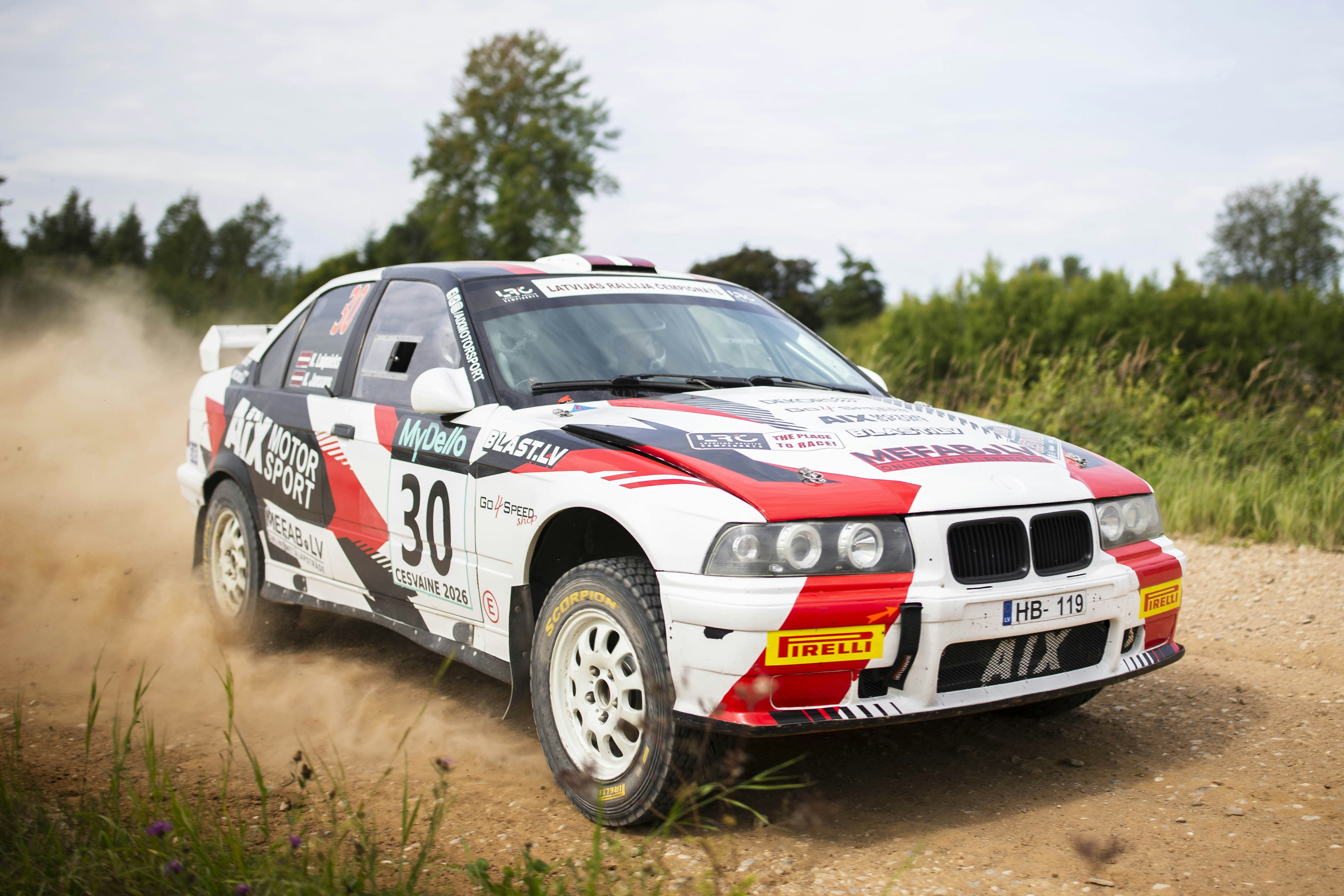 Rally car kicks up dust on a gravel road, wheels churned, sponsor graphics and number 30 visible against a rural backdrop.