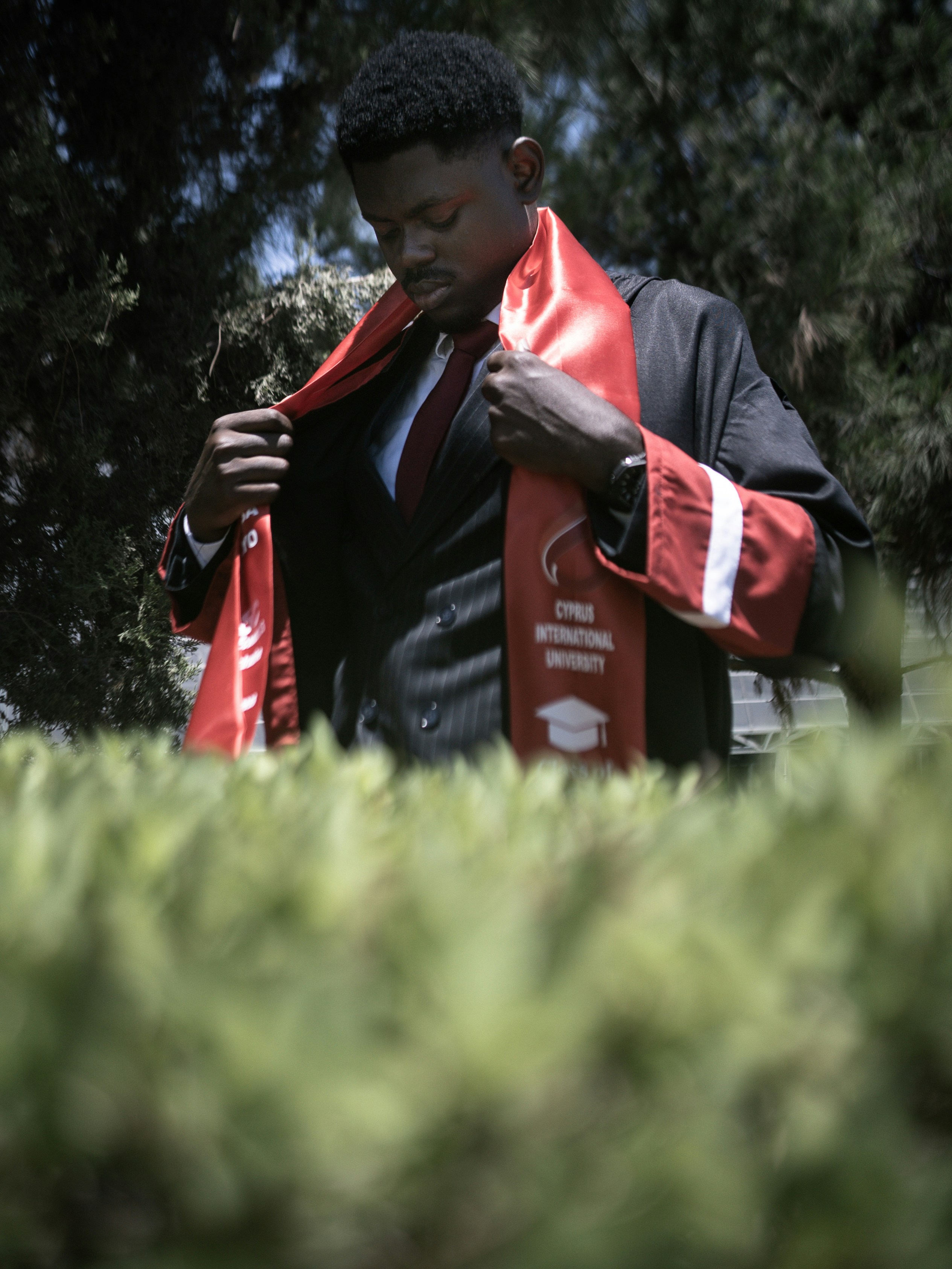 A man in a graduation gown and red sash outdoors among green foliage