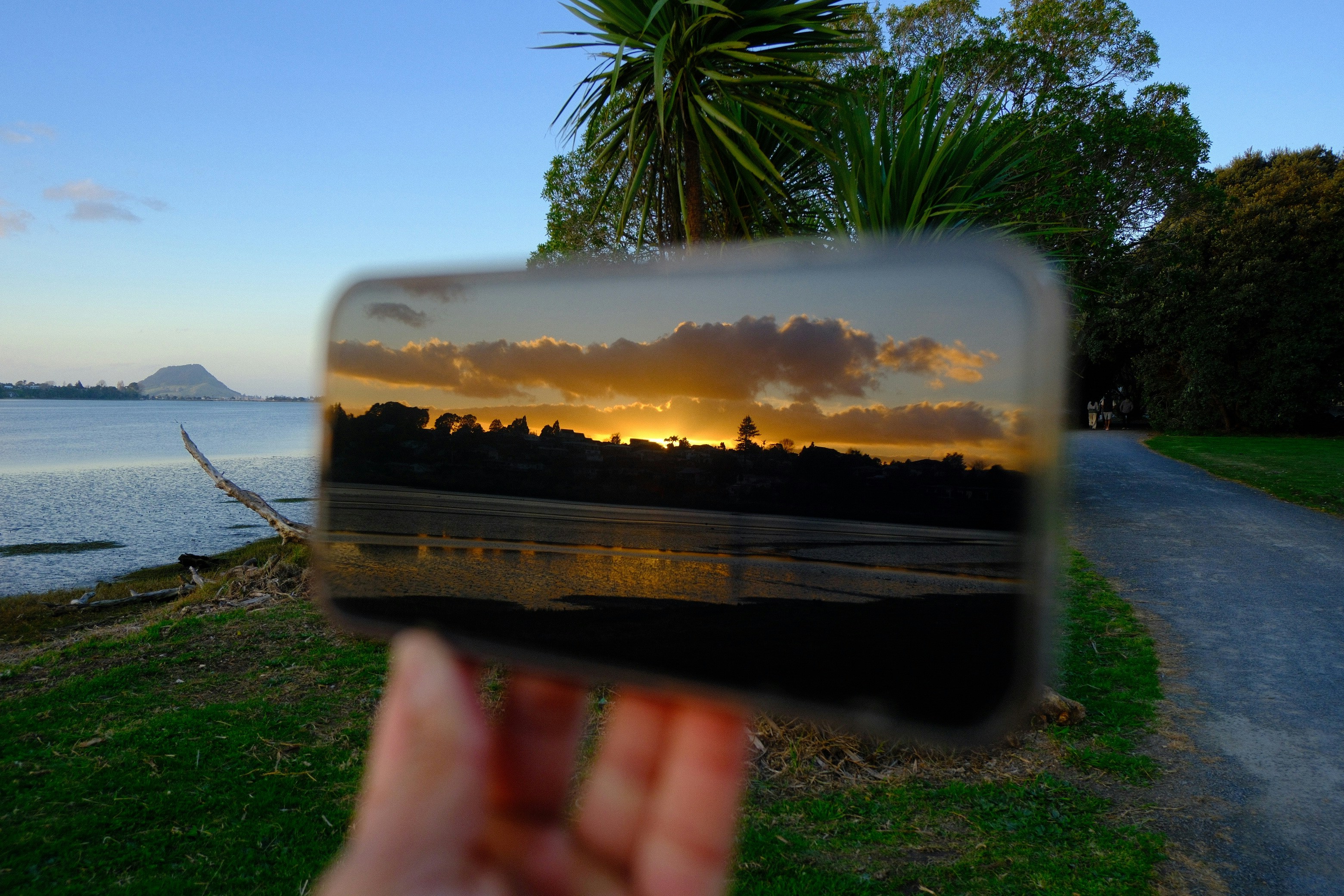 A hand-held smartphone screen reflects a sunset over a distant horizon, with the foreground and park path providing context and the lake calm behind. The scene doubles as a subtle selfie-like focal point.