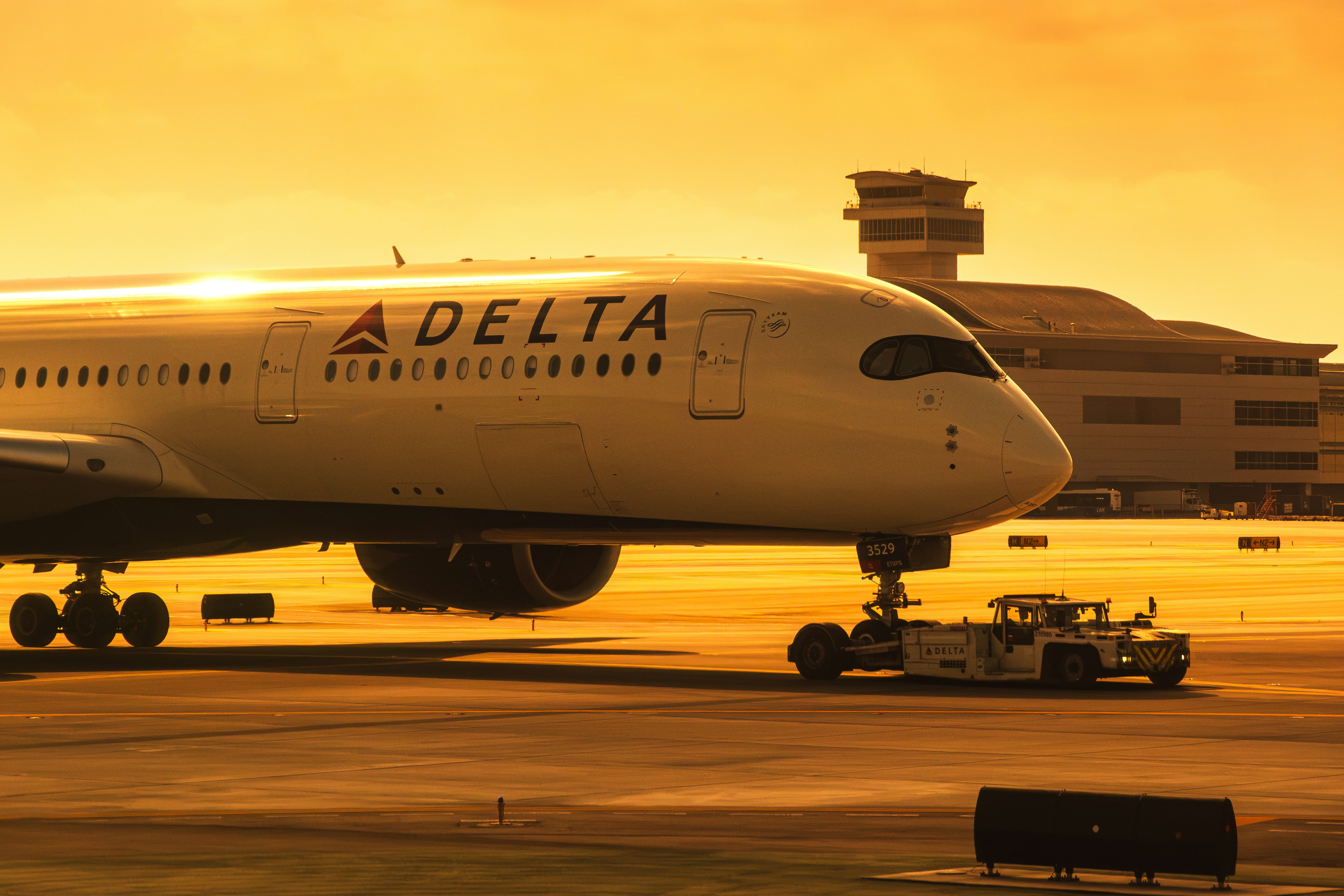 Golden-hued Delta aircraft parked on the tarmac during sunset, with ground service equipment in the foreground and a modern terminal in the background.