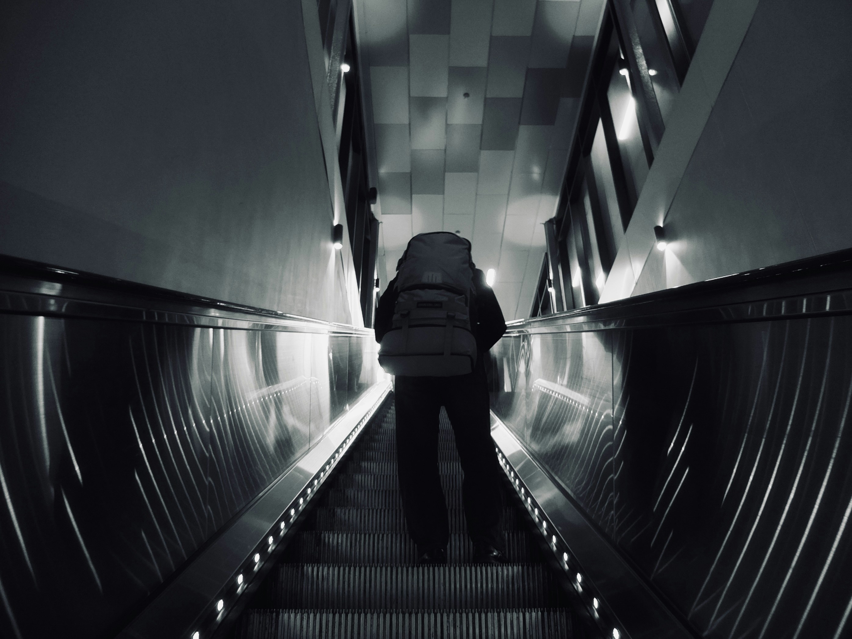 Ascend toward the Neon Quiet: A lone traveler in a monochrome escalator