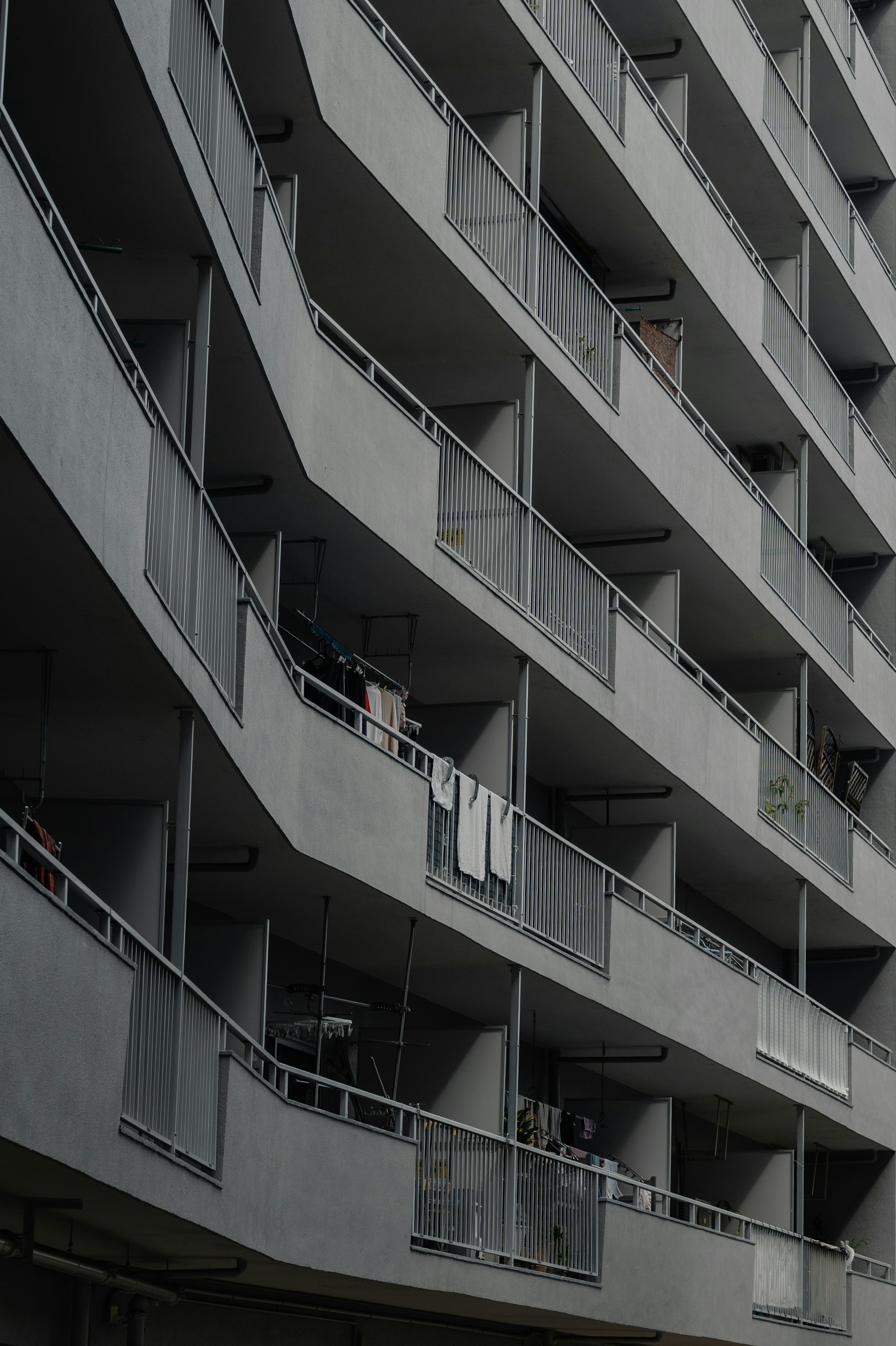 Grey concrete apartment balconies stack in diagonal rows, with laundry and plants visible on some rails, creating a repetitive architectural pattern.