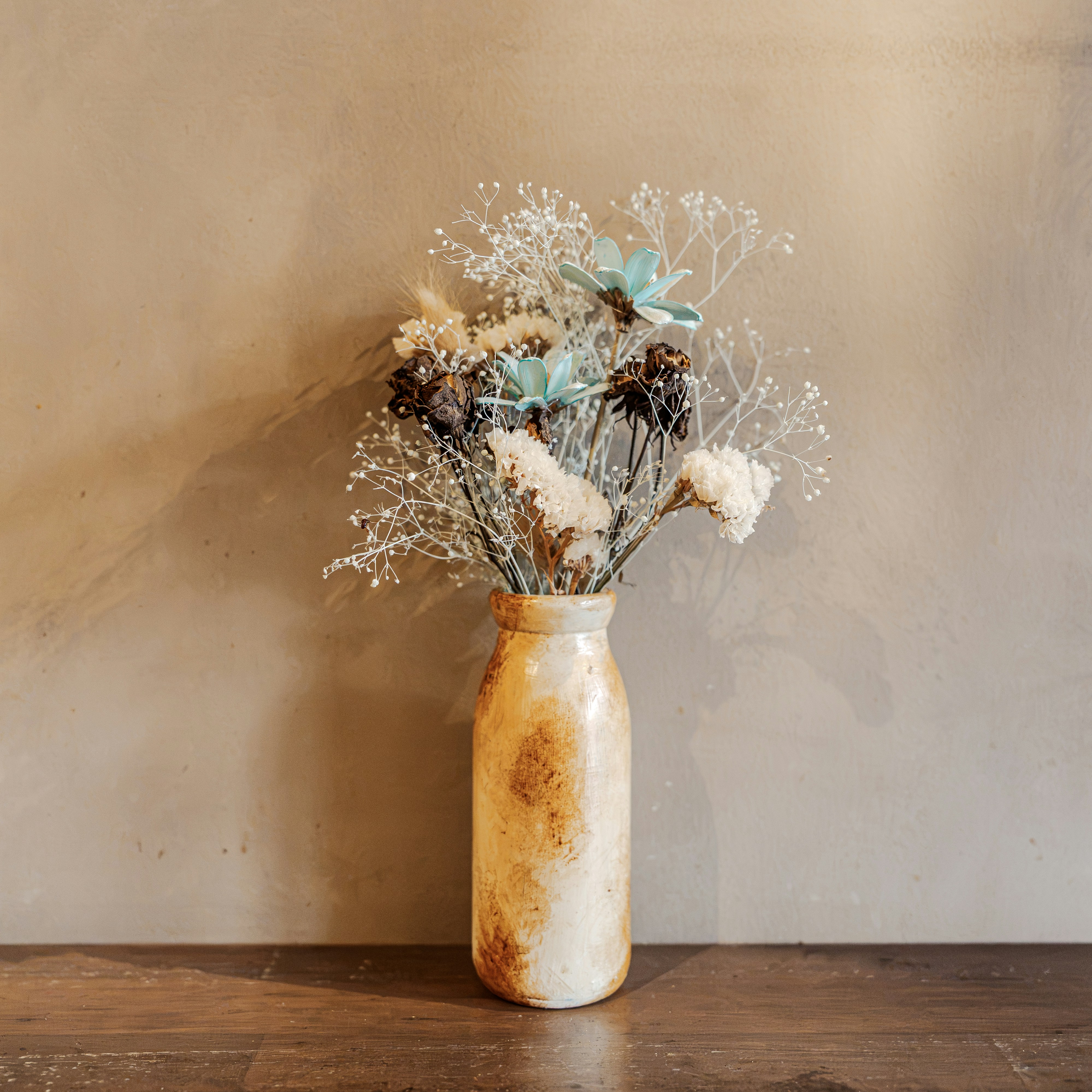 A rustic ceramic vase with dried blooms sits on a dark wooden surface against a muted wall, evoking simple, timeless still life.