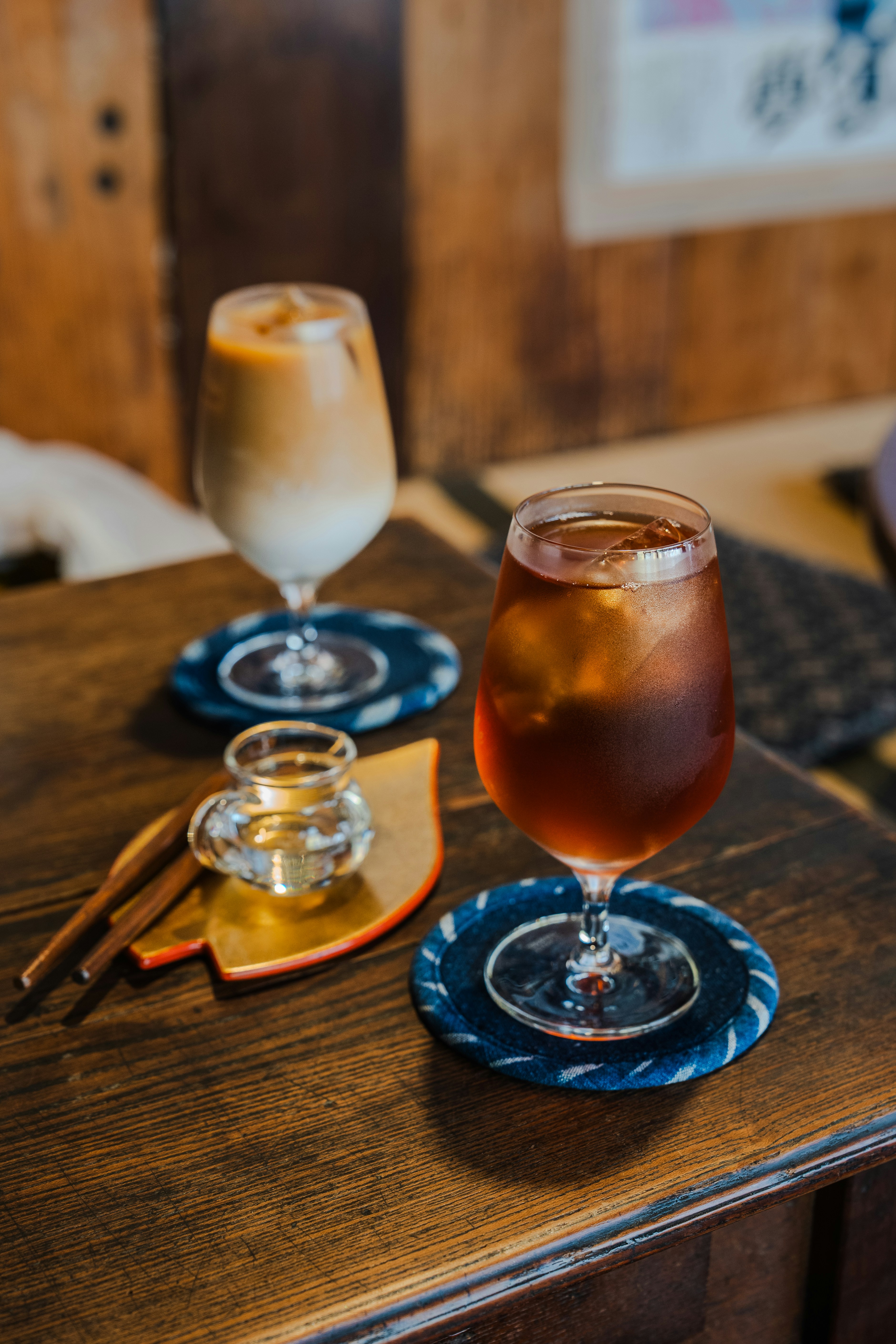 Two beverages on a wooden bar with coasters and a small glass pitcher, warm lighting and blurred background conveying a casual, intimate drink setting.