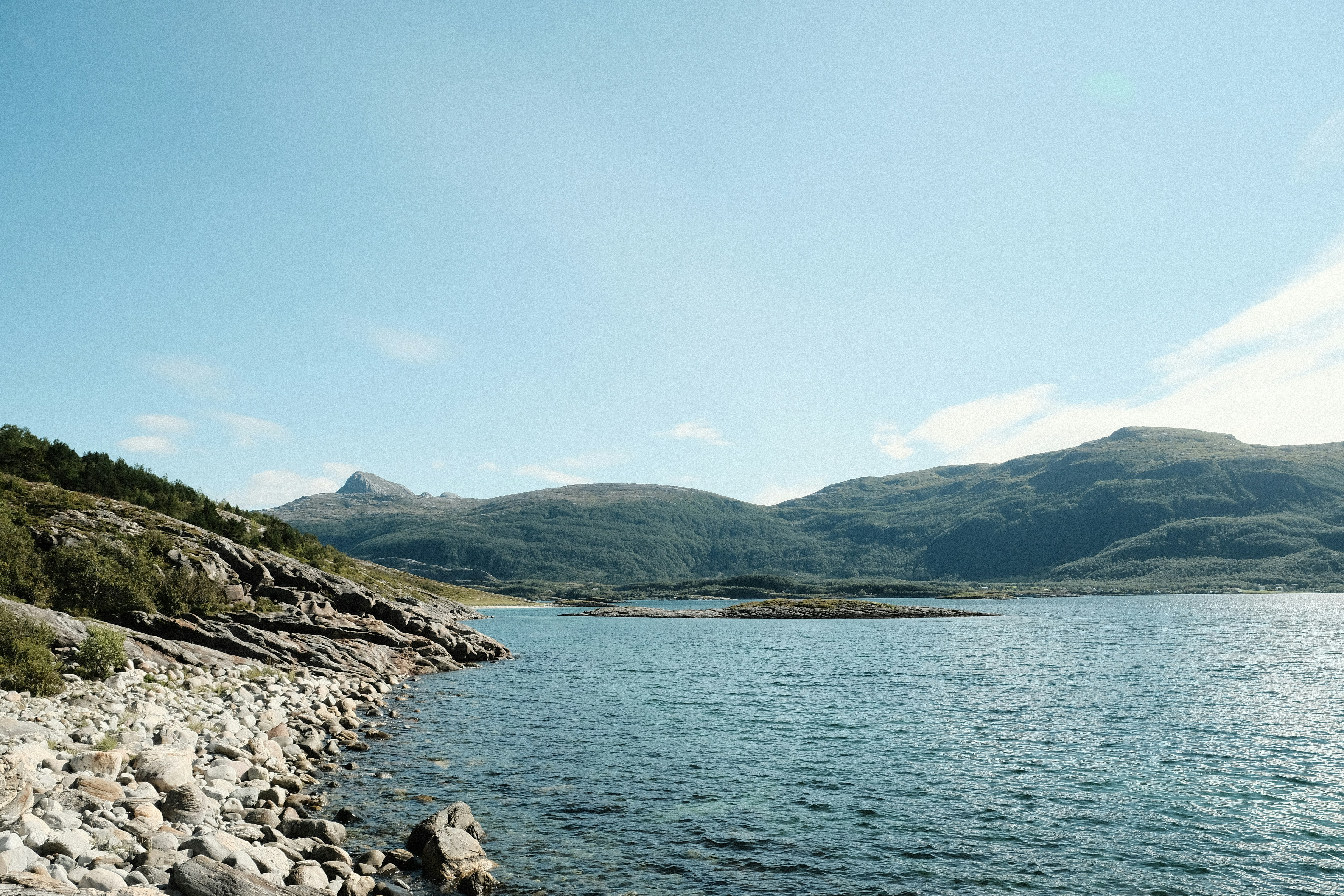 Coastal rocks meet a calm inlet with distant hills under a clear blue sky; the scene emphasizes rugged shoreline leading into tranquil water.