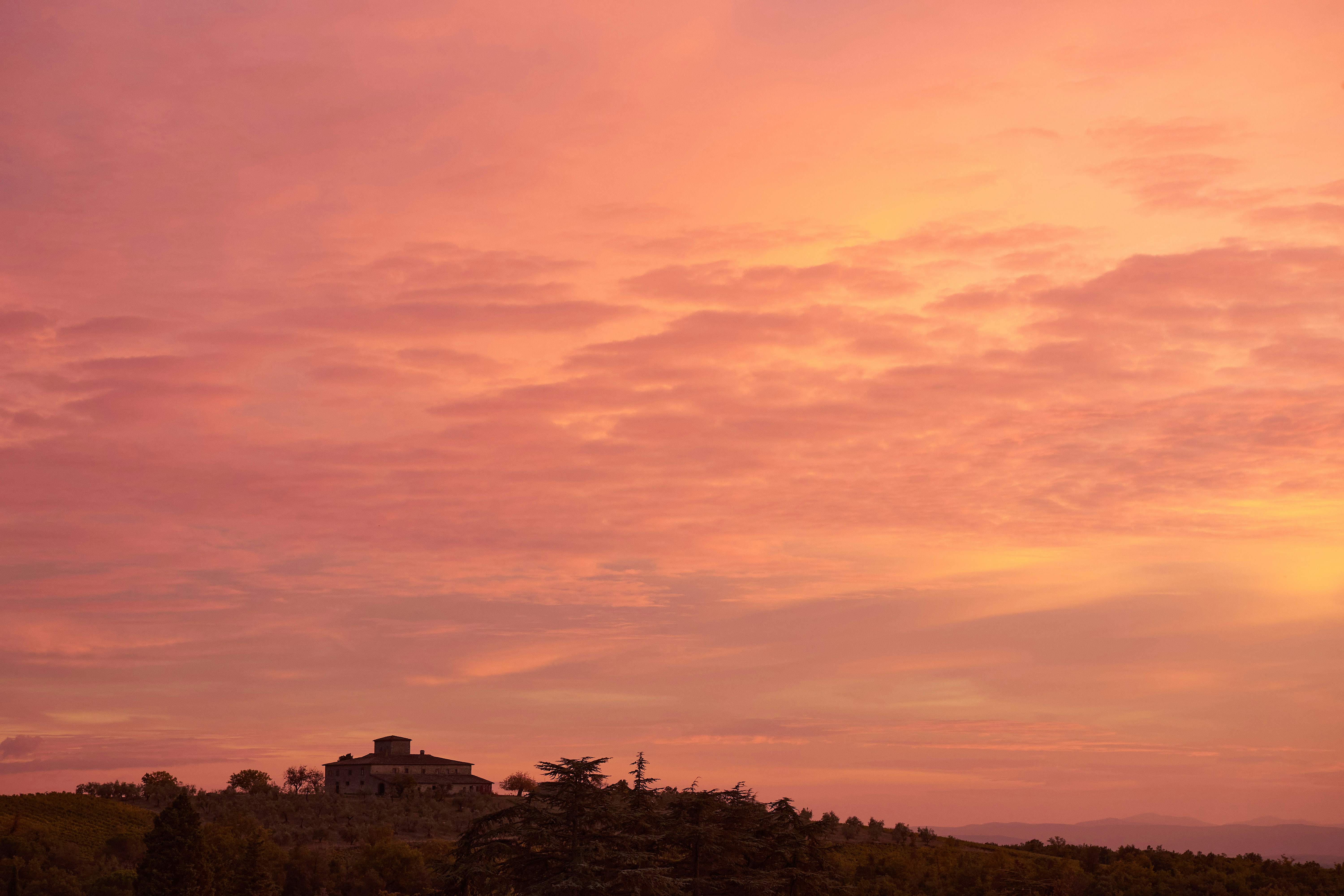 Silhouetted hill and building beneath a warm, cloud-streaked sunset sky with soft pastel tones.
