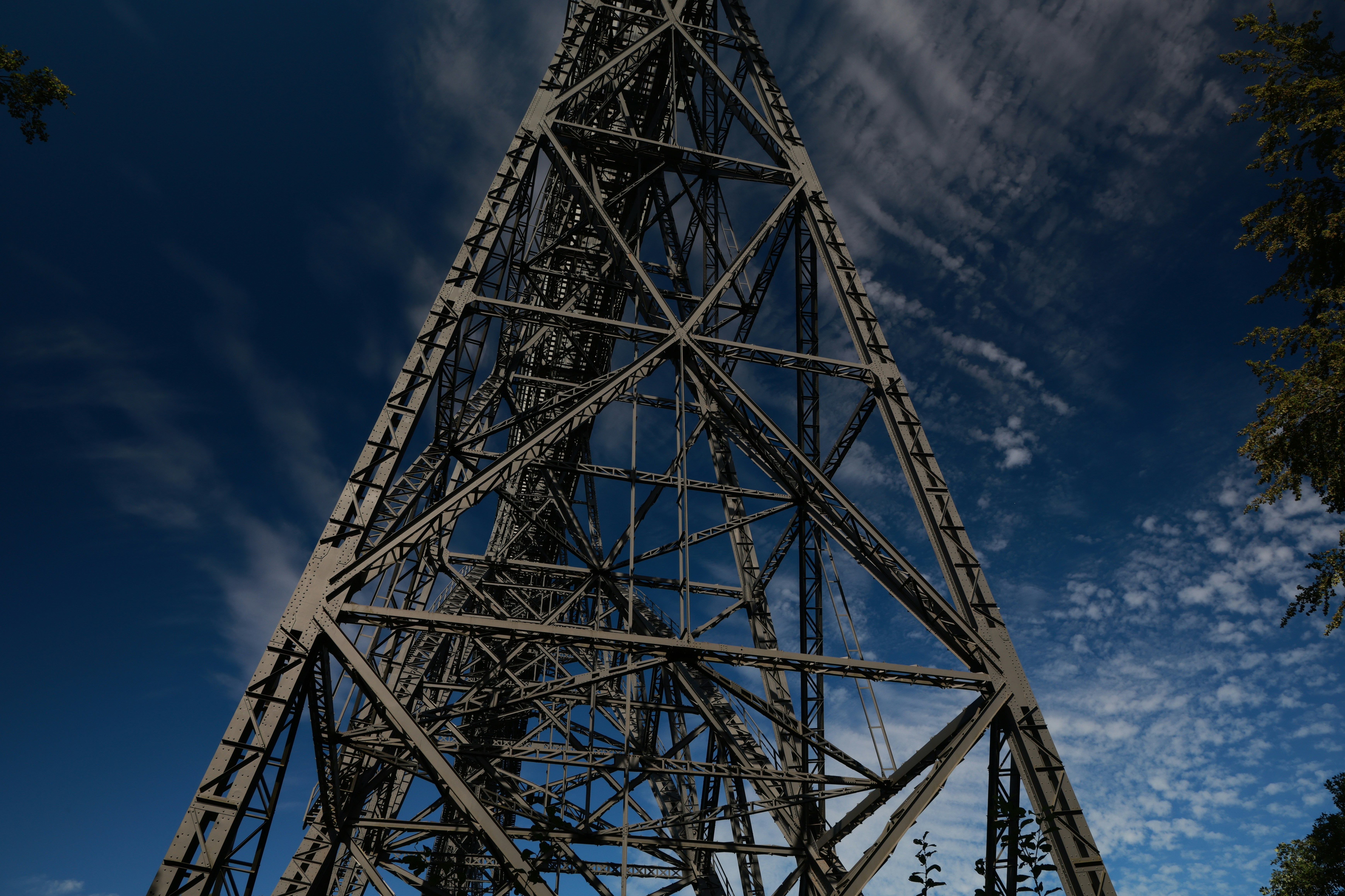 Industrial lattice tower looms against a deep blue sky with wispy clouds, captured from a low angle.