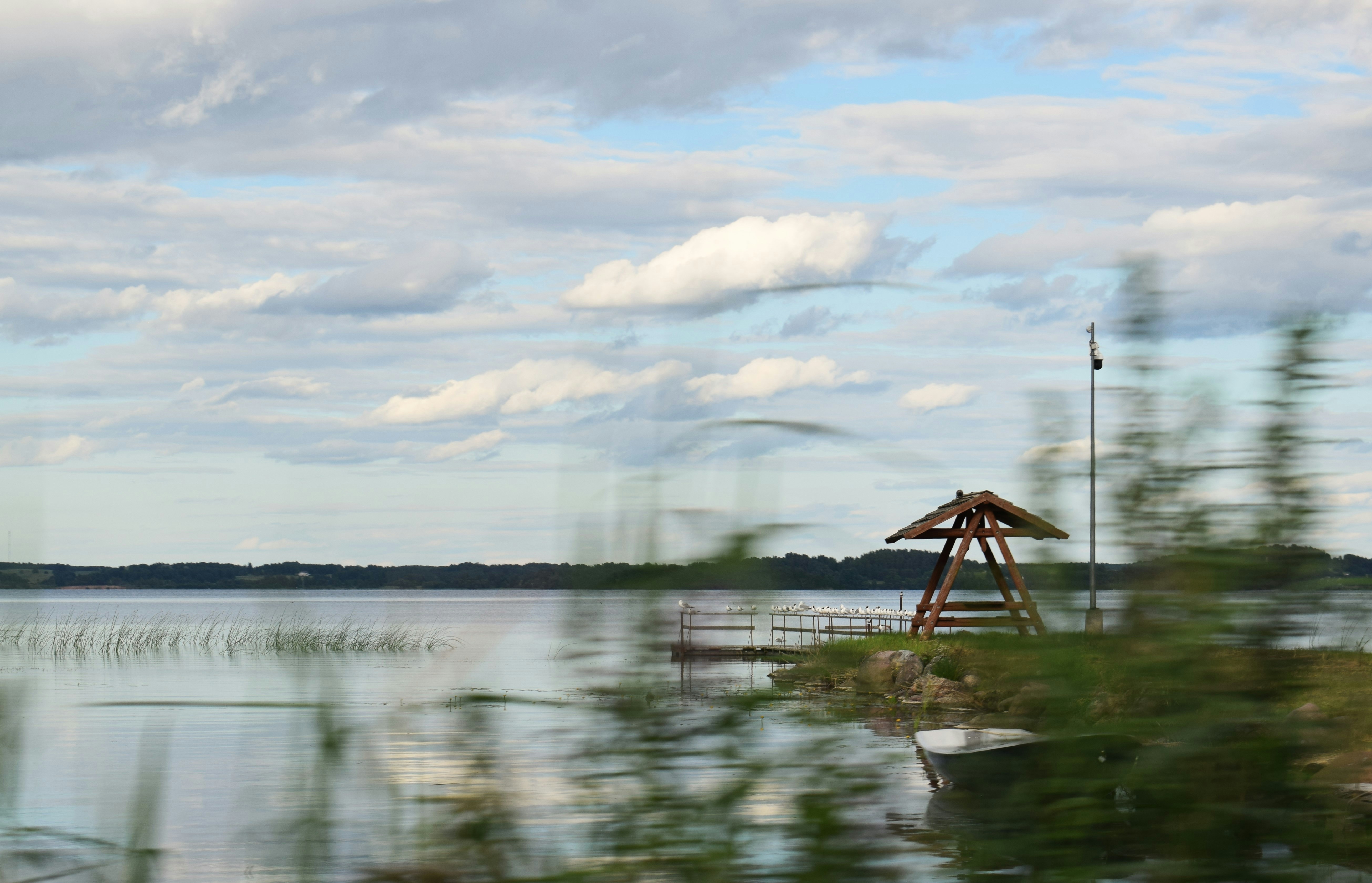 Blurred lakeside scene with a wooden gazebo and pier by the water; foreground grasses are out of focus, suggesting motion.