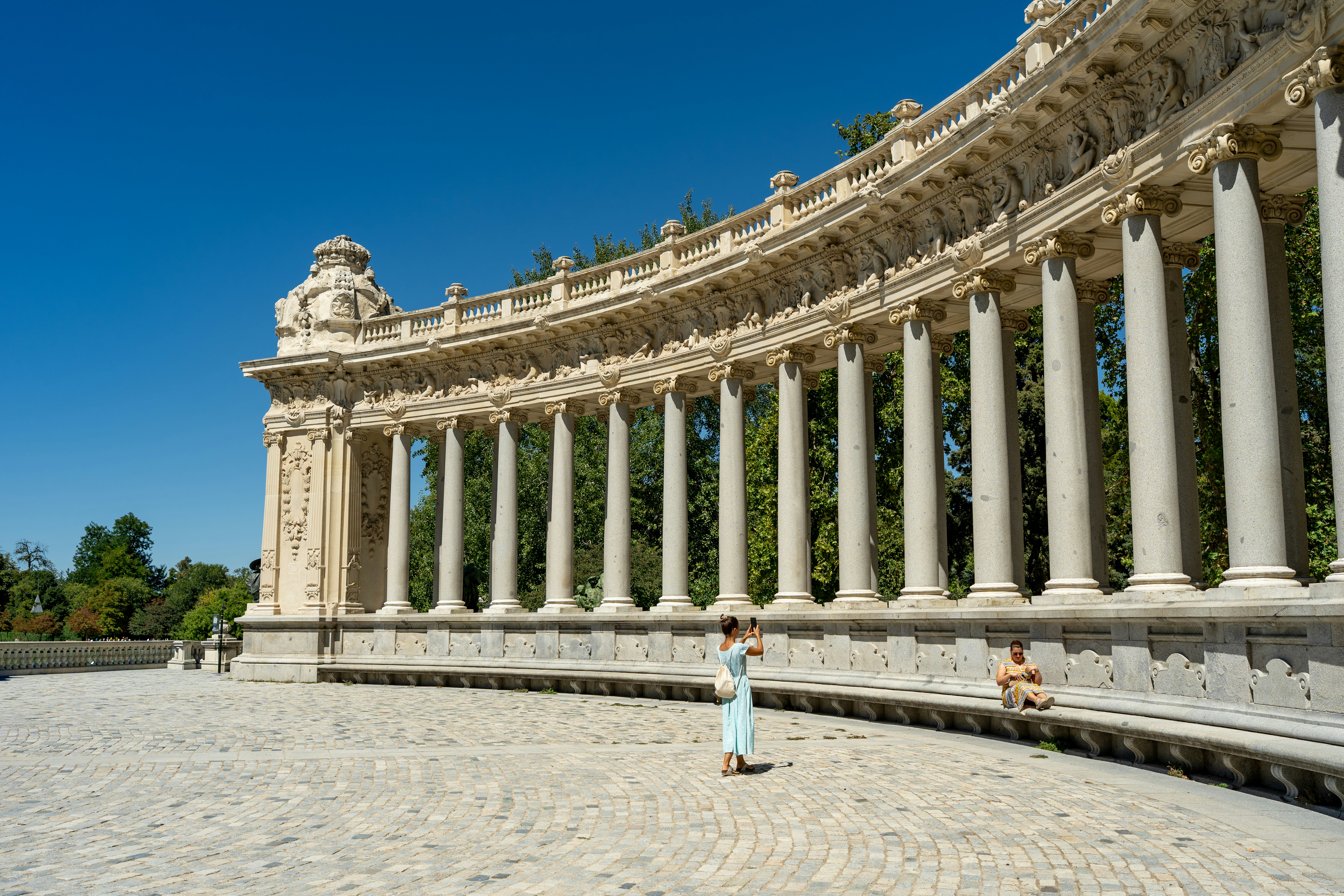 Wide-angle view of a curving stone colonnade with tall columns, a cobblestone plaza, and two visitors—one center foreground and another seated—under a clear blue sky.