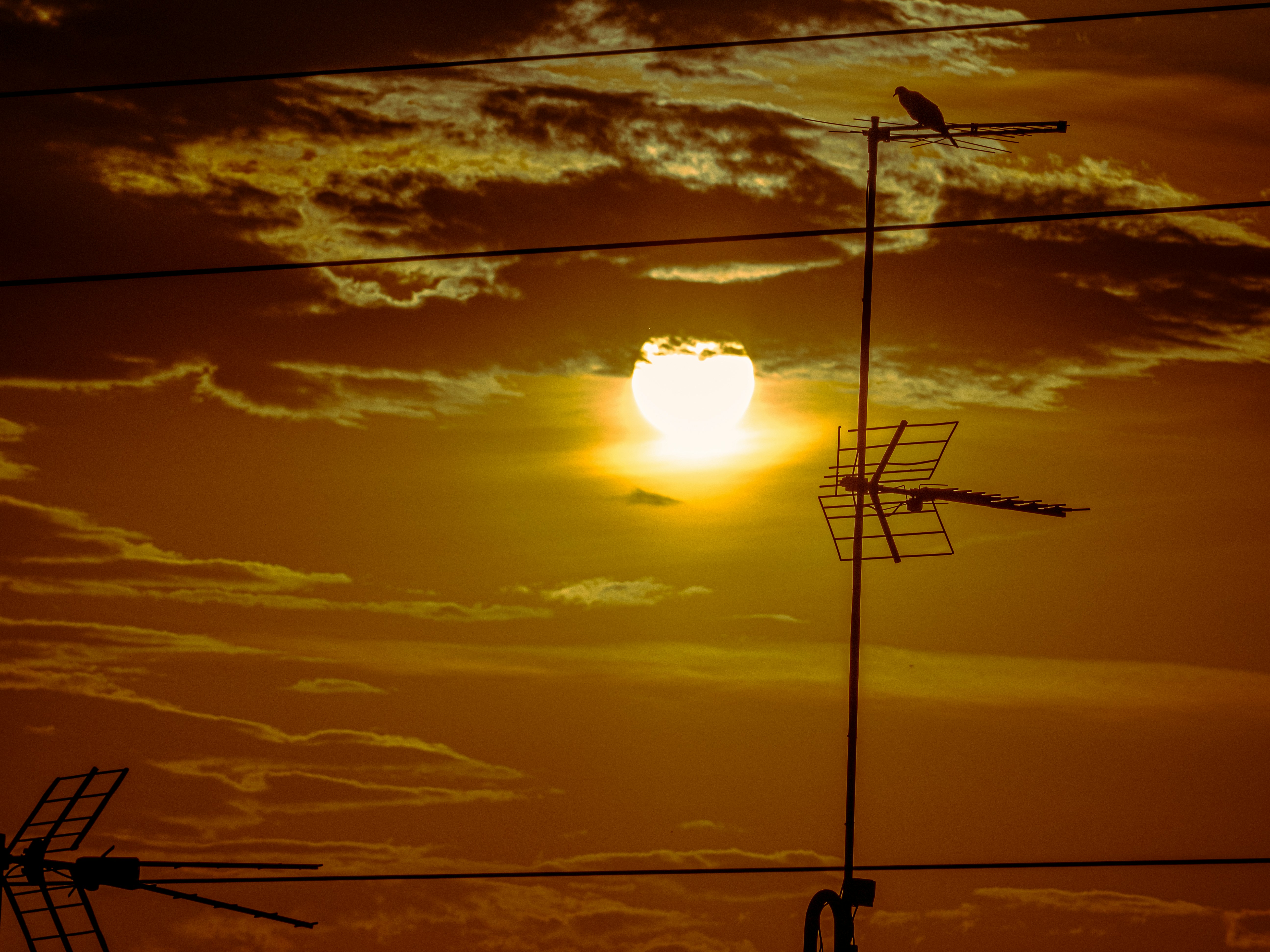 Rural rooftop antennas silhouette against a vivid orange sunset sky with scattered clouds.