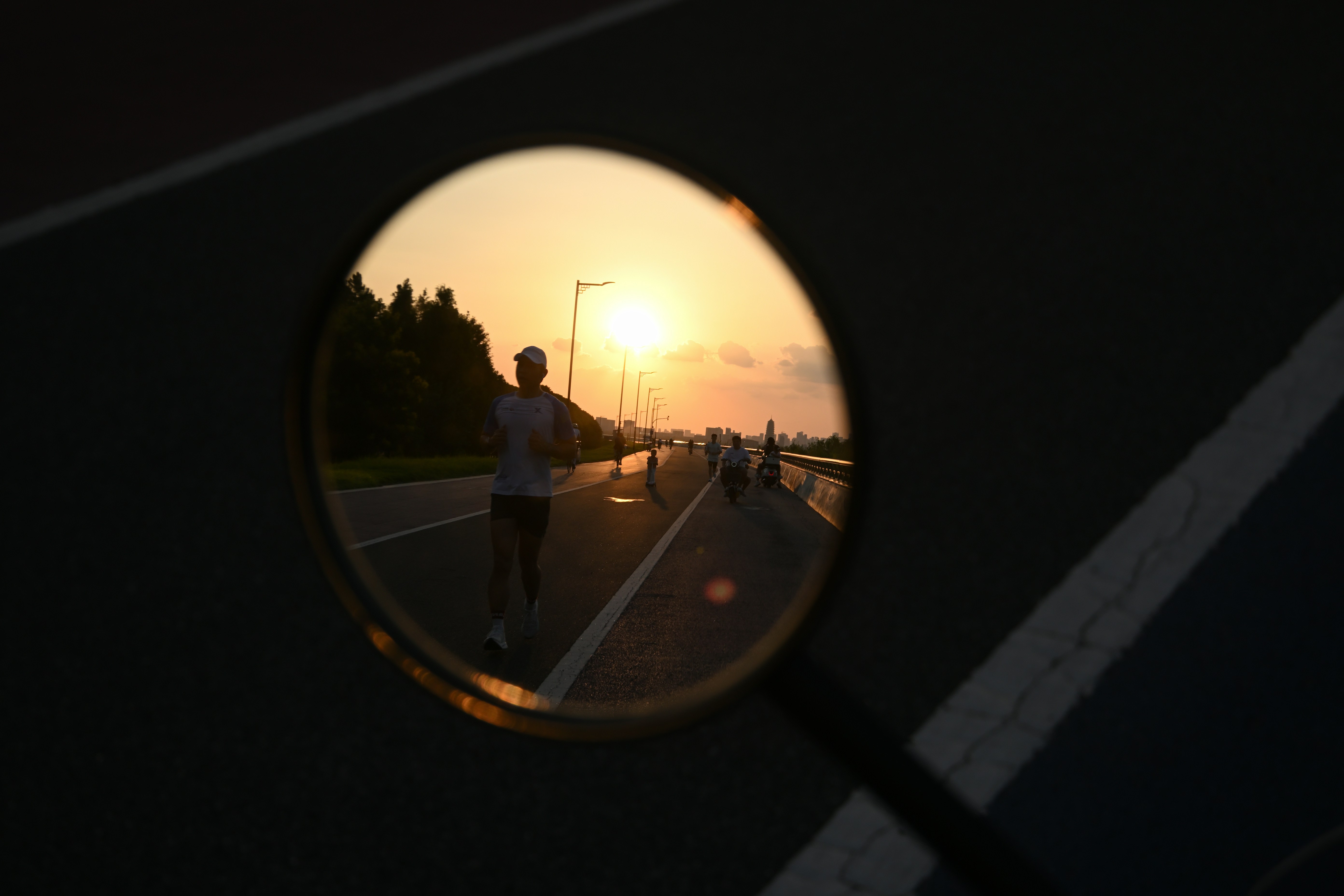 Runner silhouette captured in a circular reflection on a roadside mirror at sunset, with distant cyclists and a warm sky. A logo-like circular frame emphasizes motion and urban context.