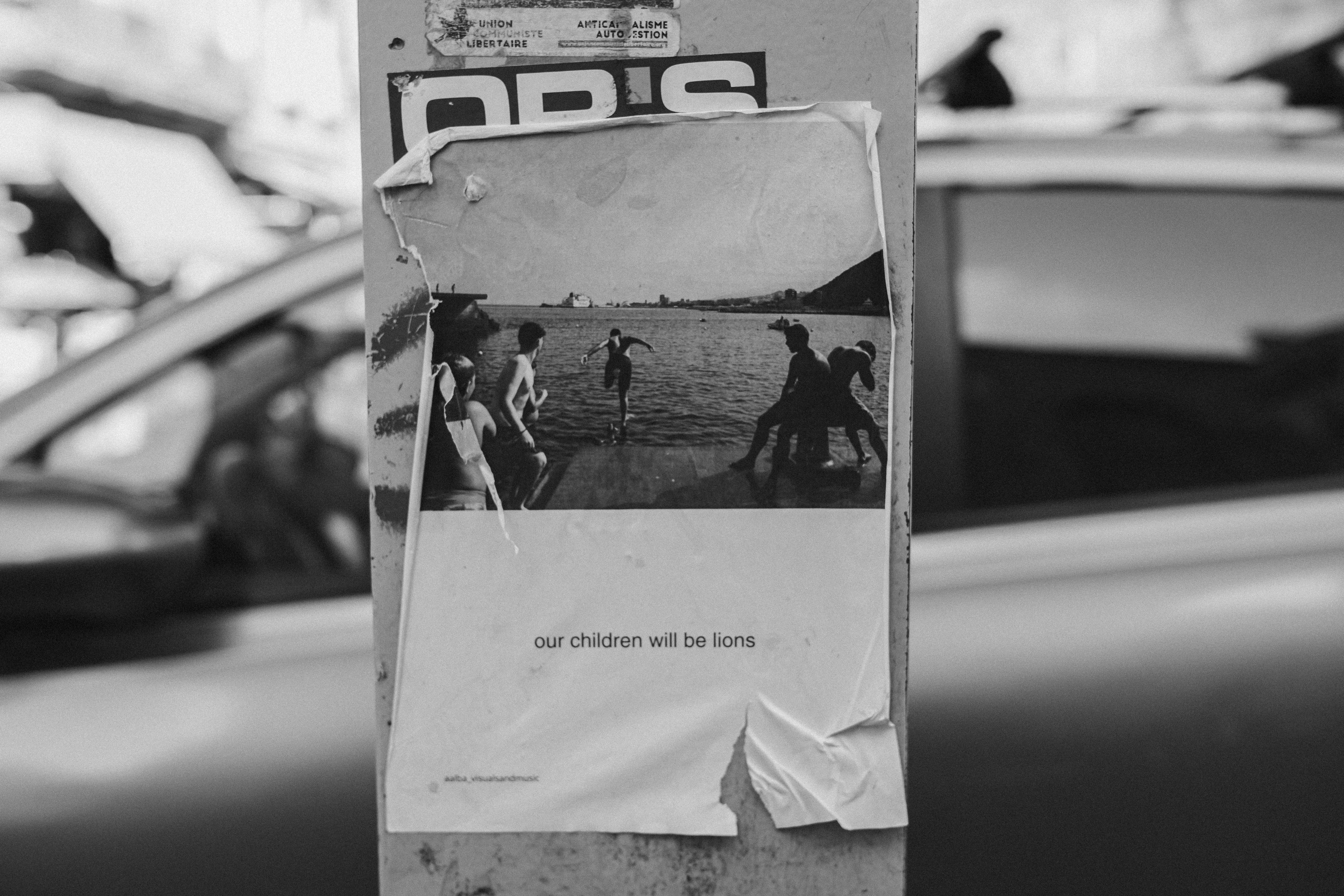 A grayscale street scene featuring a torn poster affixed to a weathered pole, with blurred vehicles in the background and a small group of people depicted on the poster itself.