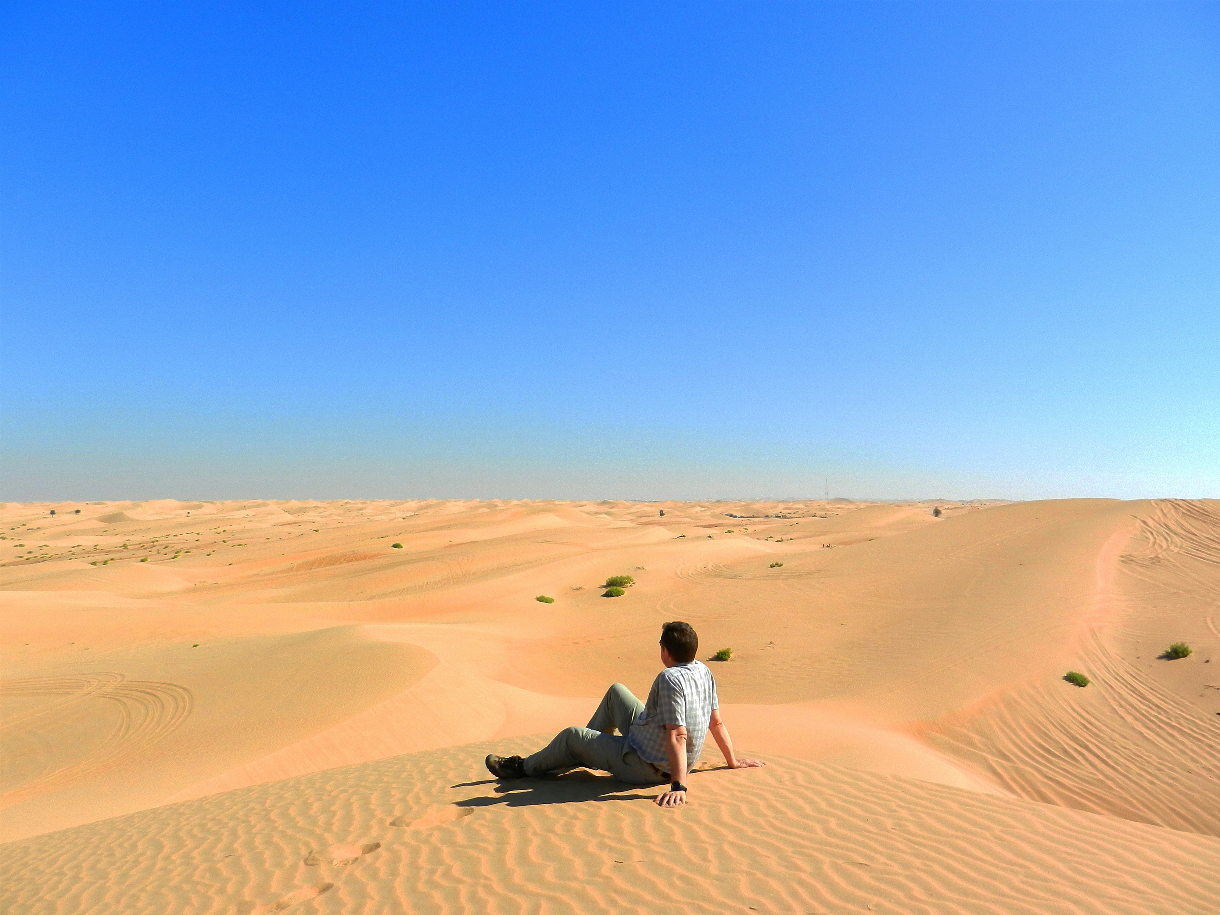 Desert dunes under a clear blue sky with a lone traveler resting on a dune crest, illustrating scale and solitude; a photographic capture of a sparse landscape.