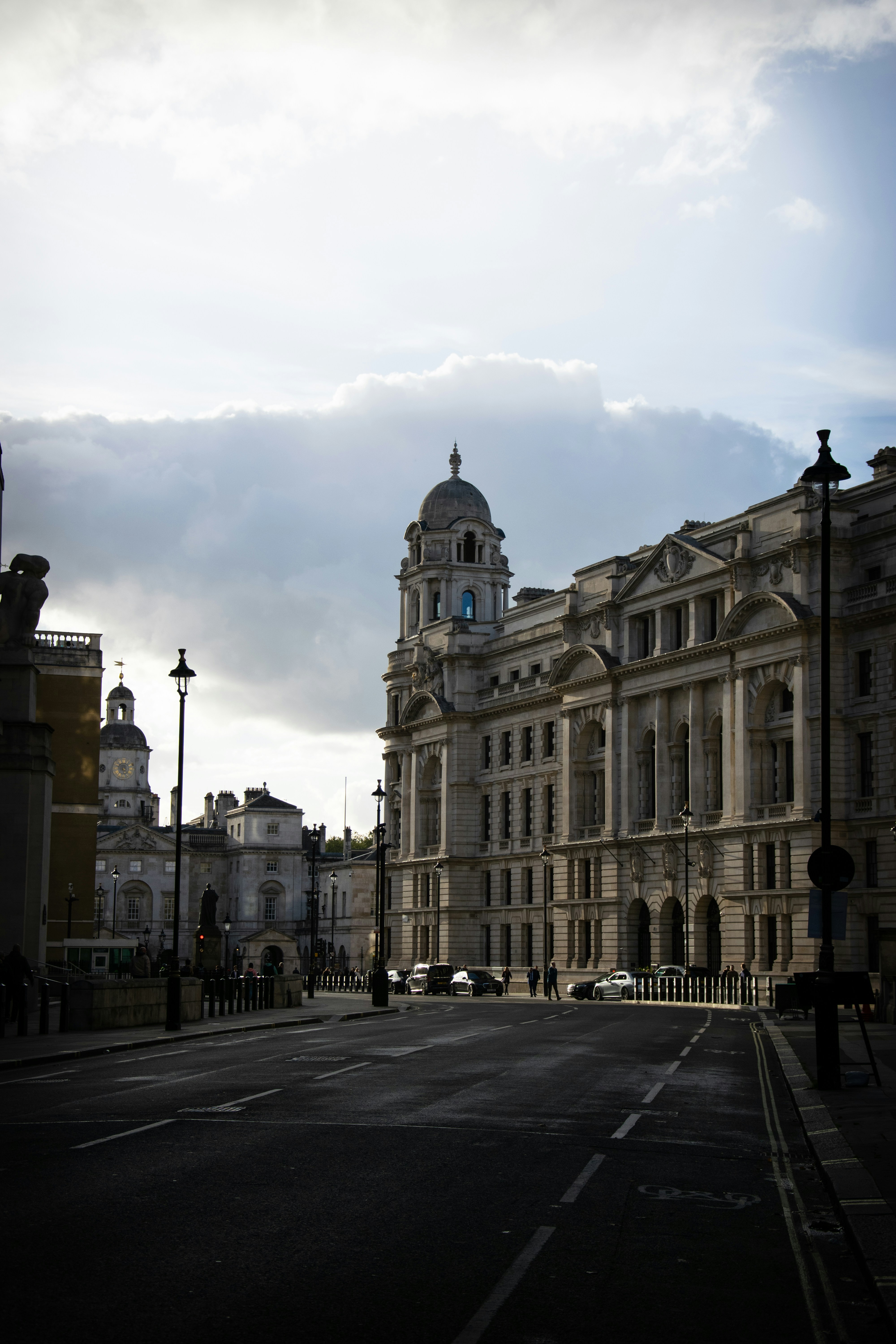 Stone neoclassical buildings line a quiet city street, with a grand central facade and a clock tower set against a partly cloudy sky.