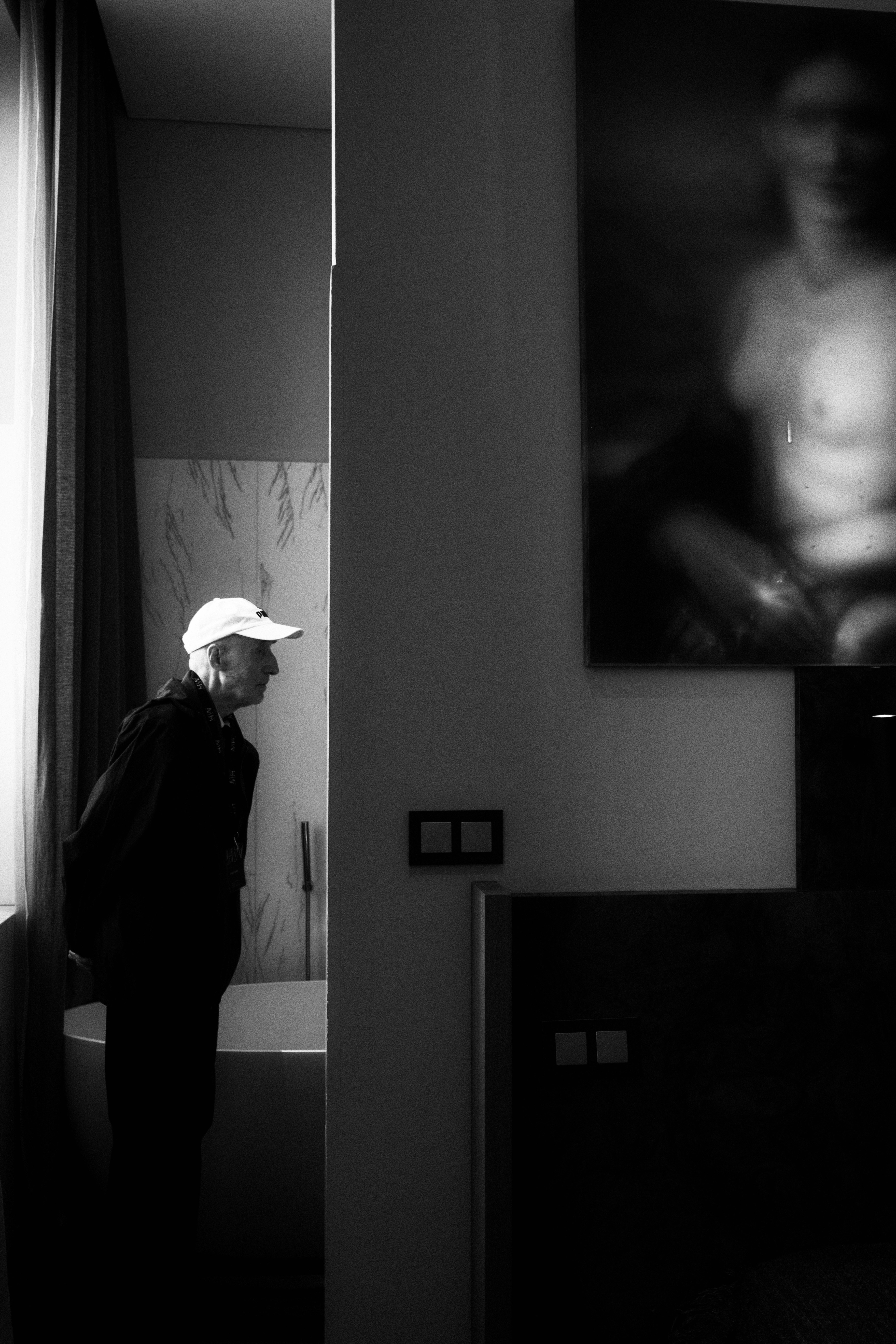 Older man in a white cap stands near a doorway in a dim, monochrome interior with art on the wall to the right.
