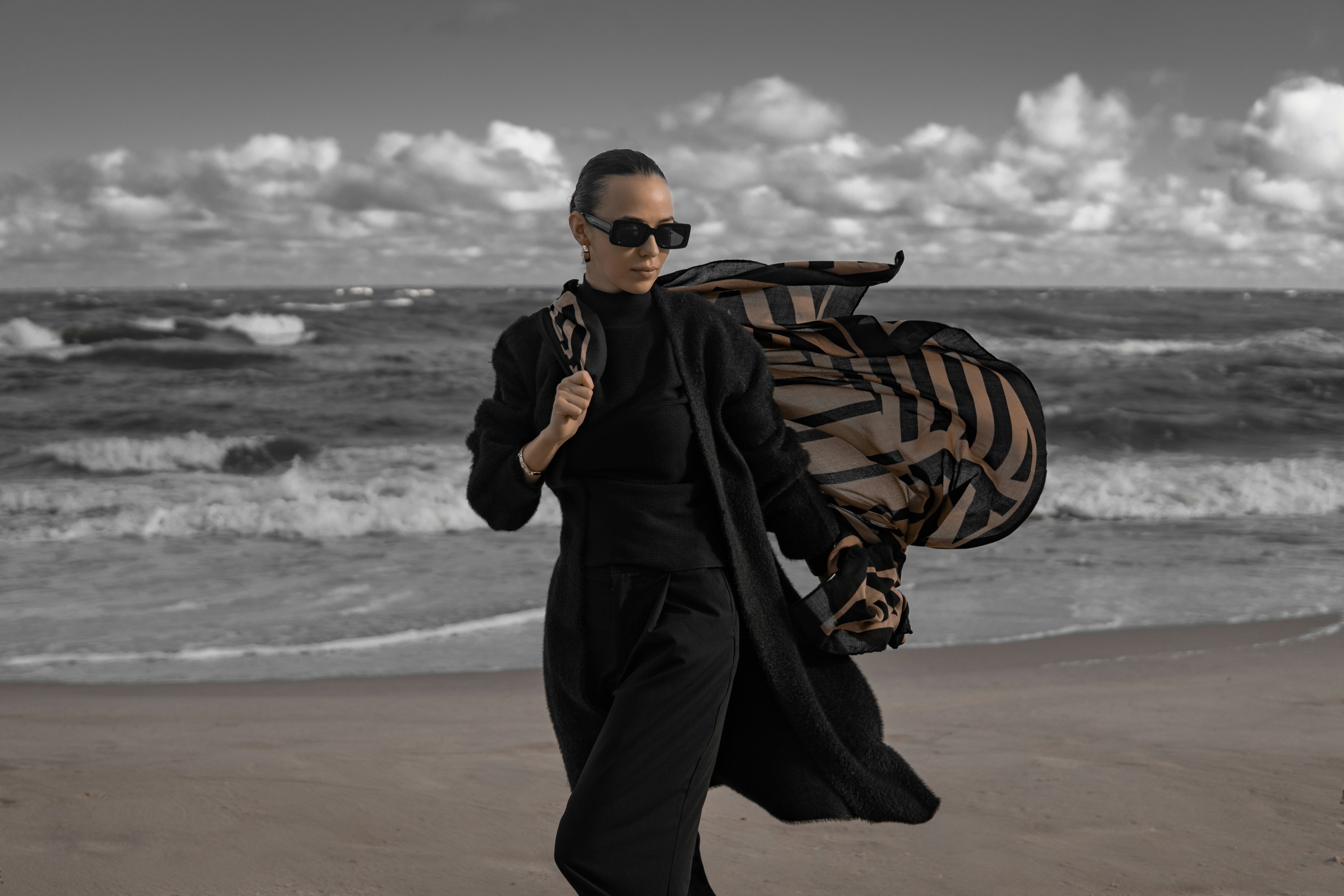 A stylish figure in black, sash and sunglasses stands on a windy beach with a patterned scarf billowing behind them, creating a dramatic portrait against the ocean backdrop.