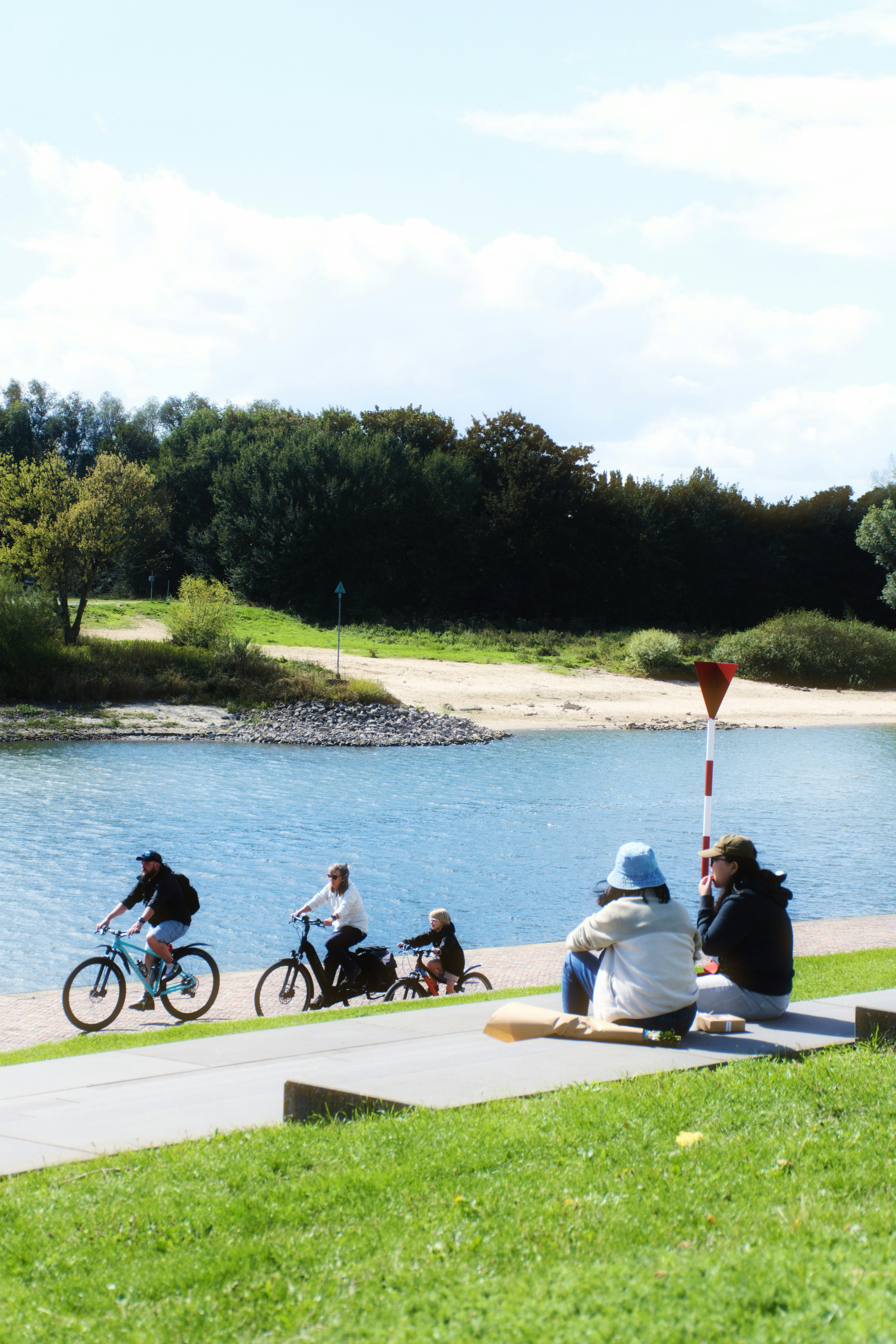 A group enjoys a sunny day along a riverside walkway with cyclists passing by and two people seated on a bench by the water.