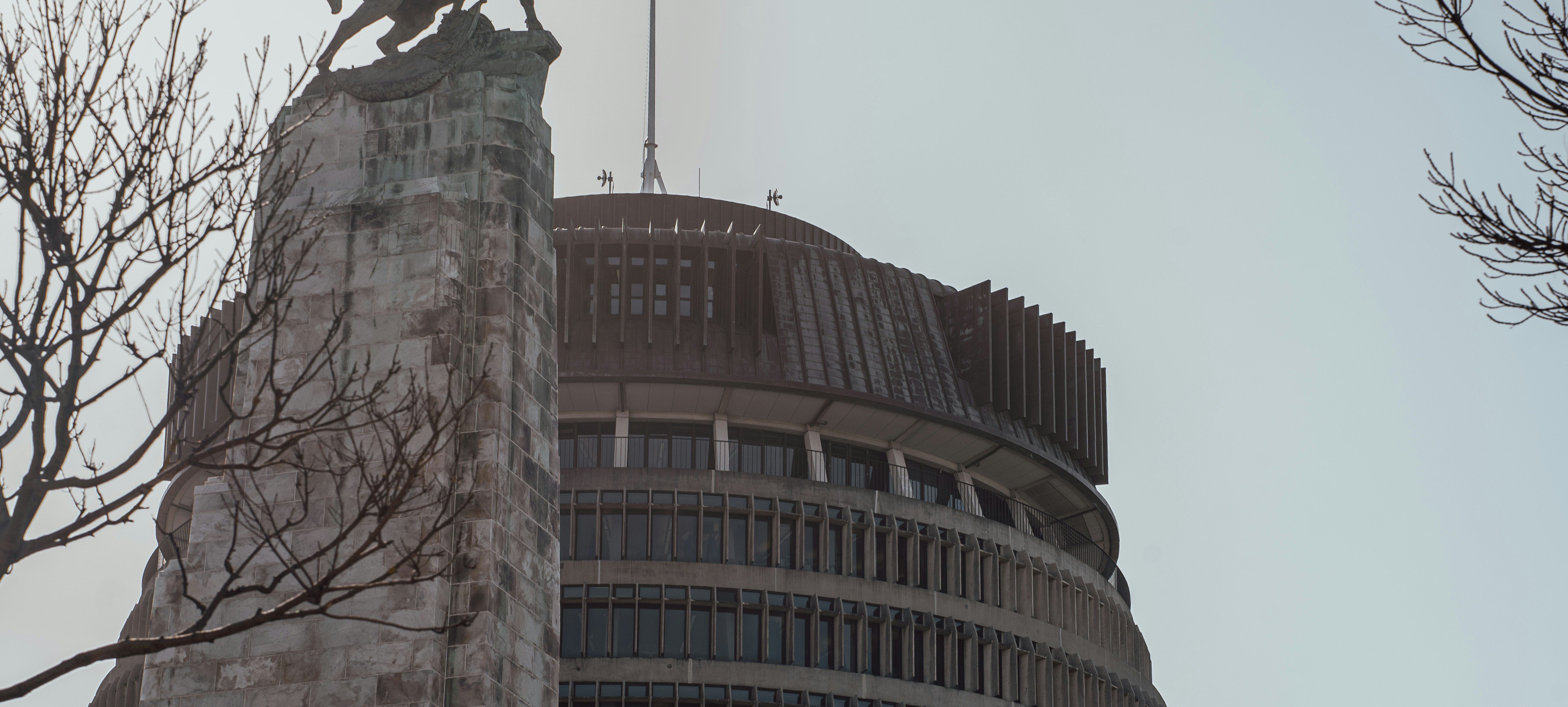 Architectural detail of the Beehive beside a stone monument and bare tree branches in Wellington, New Zealand.