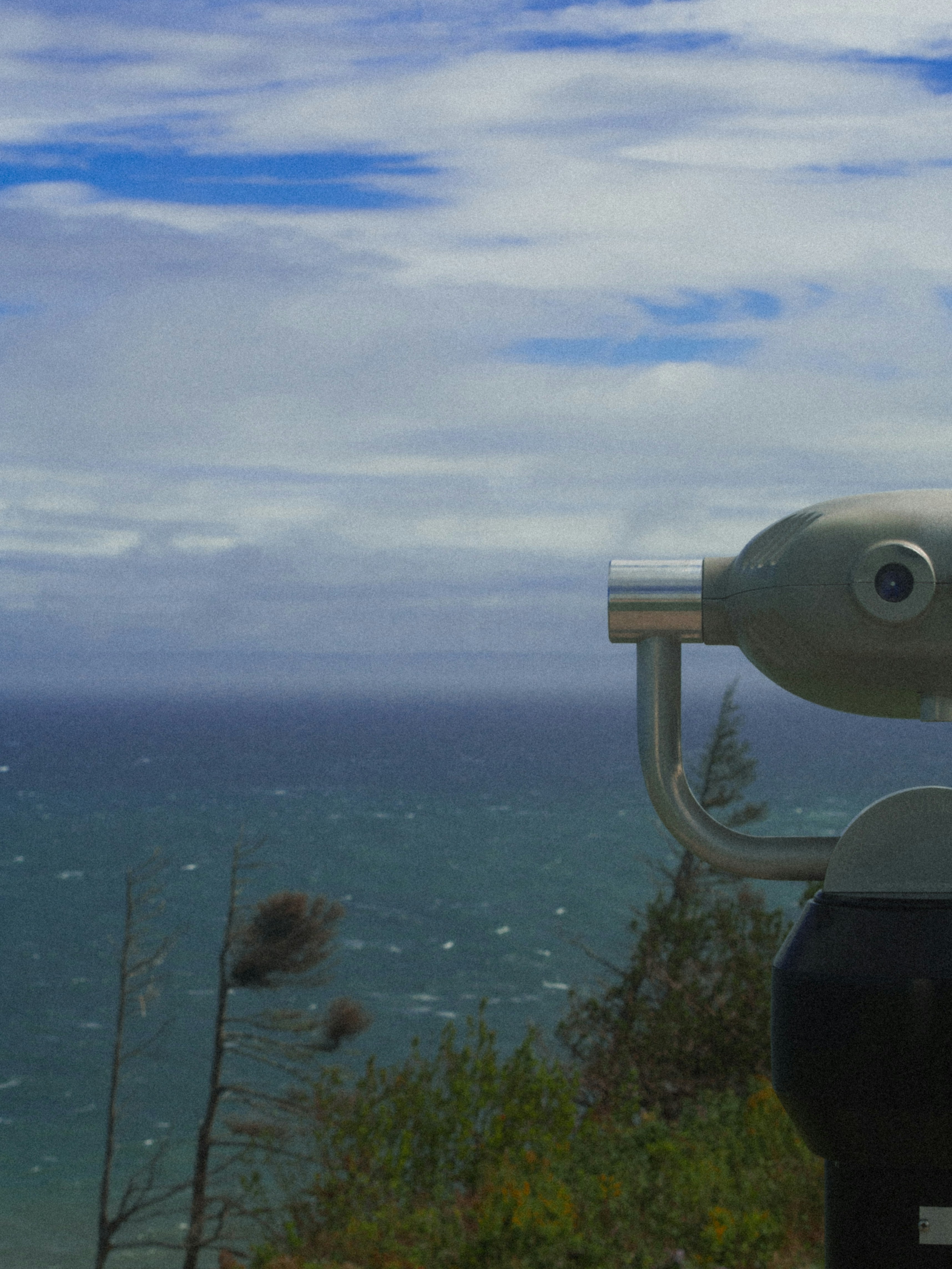Coastal horizon with a mounted viewer pointing toward the sea; foreground shows vegetation and a distant expanse of ocean and sky. This is a photograph captured with a scope-like device in the frame.
