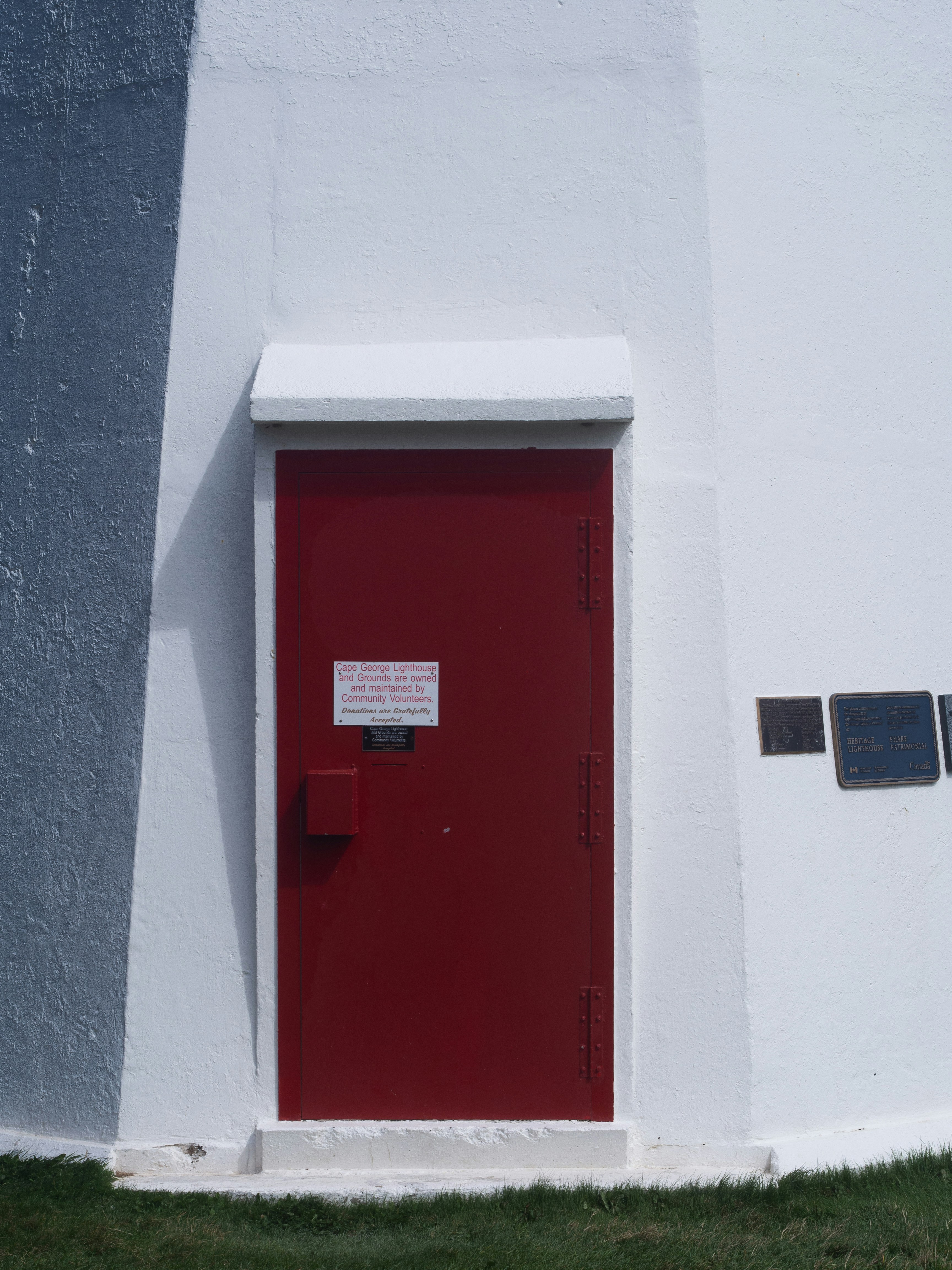 A bold red door set into a stark white wall, with a small plaque and a grassy base; architectural minimalism and strong color contrast.