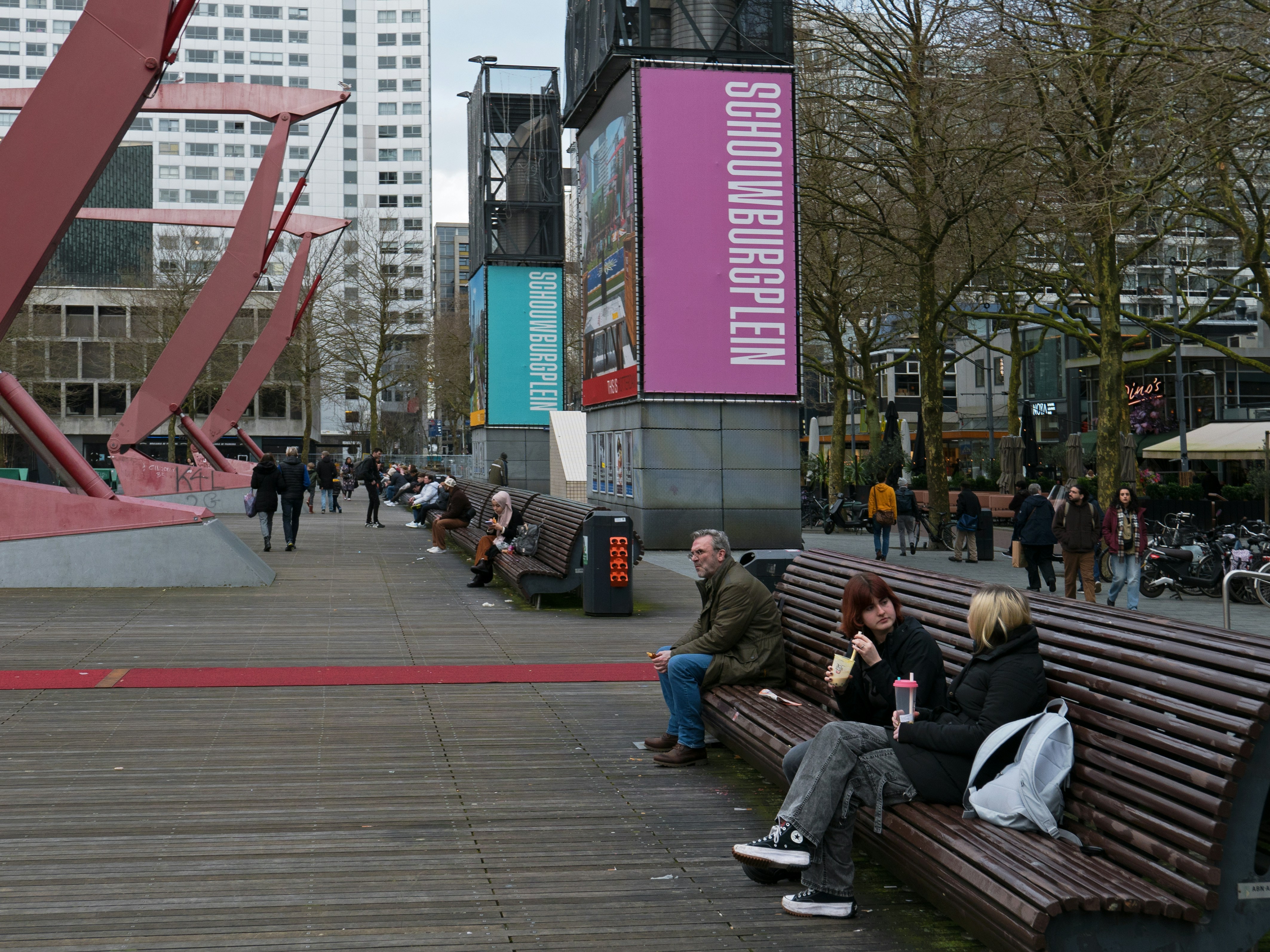 Urban Quiet: People Rest on Wooden Curved Benches by a Red Modern Sculpture