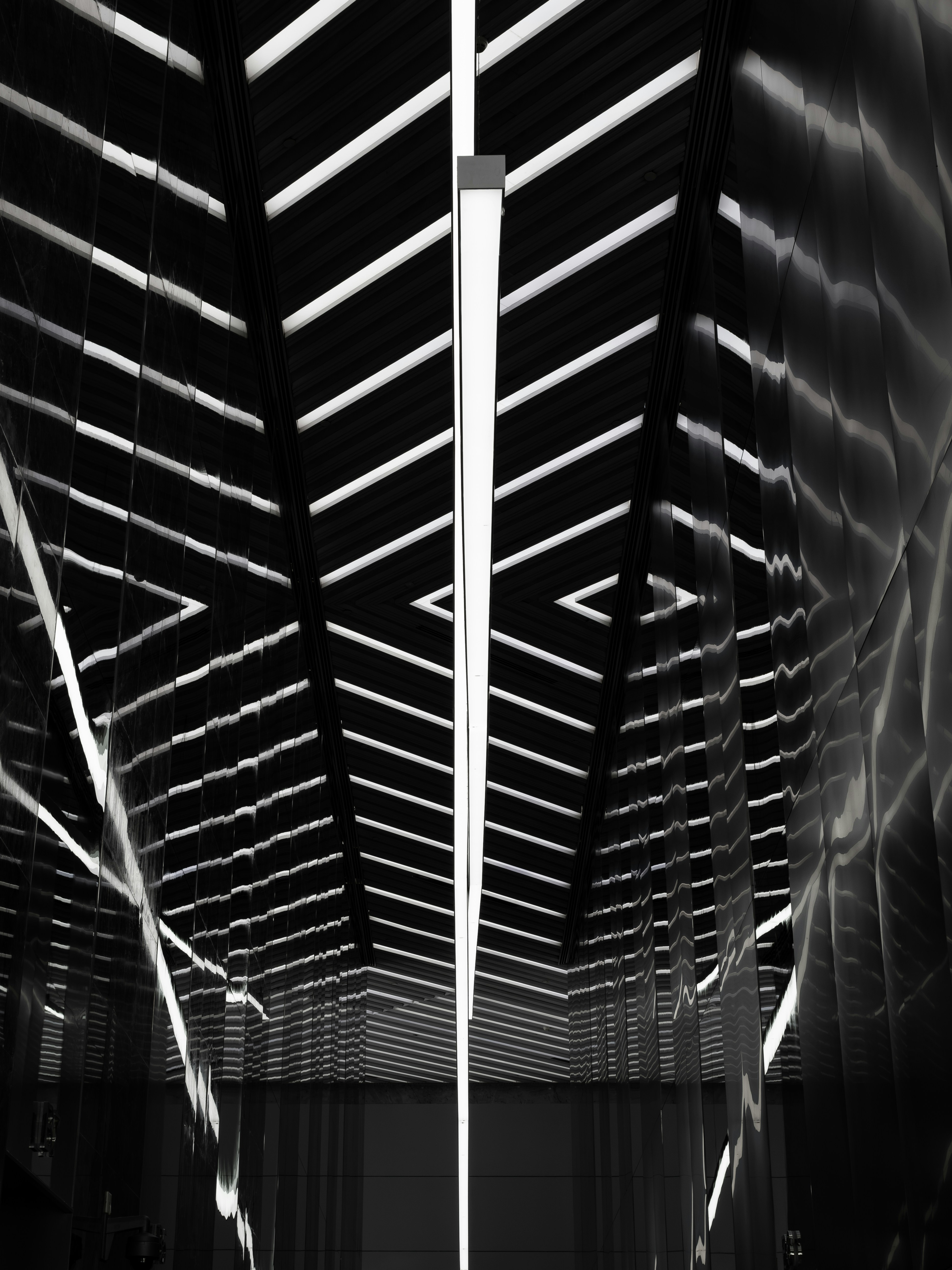Industrial black-and-white corridor with mirrored walls and a central vertical light strip, creating a geometric tunnel of repeating patterns. Abstract architectural scene with high contrast and reflective surfaces.