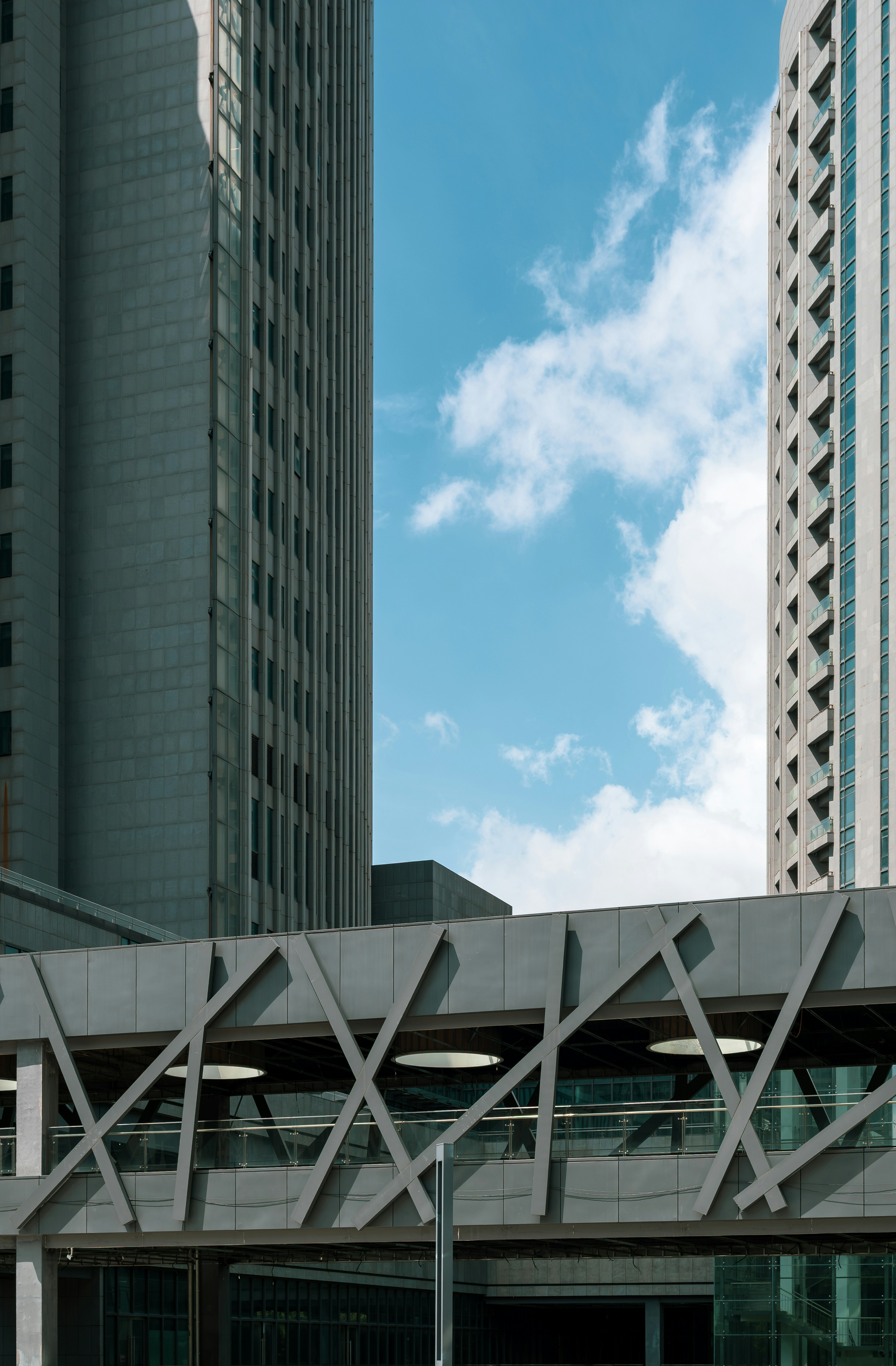 Tall, geometric buildings frame a bright blue sky with scattered clouds, while a modern pedestrian bridge introduces diagonal patterns at the lower edge.