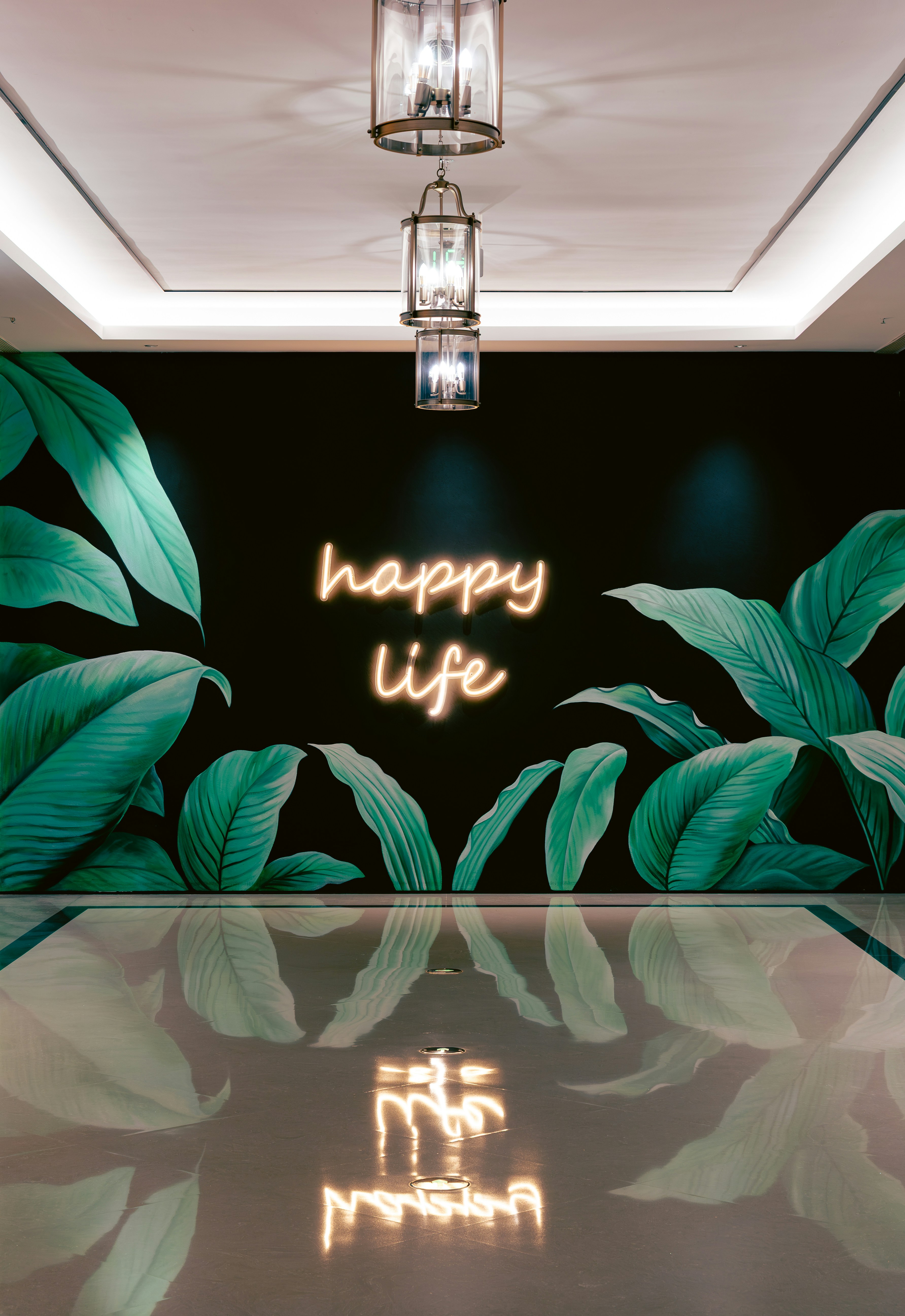 Neon 'happy life' lettering glows against a dark wall framed by tropical green leaves and a glossy floor reflecting the scene.