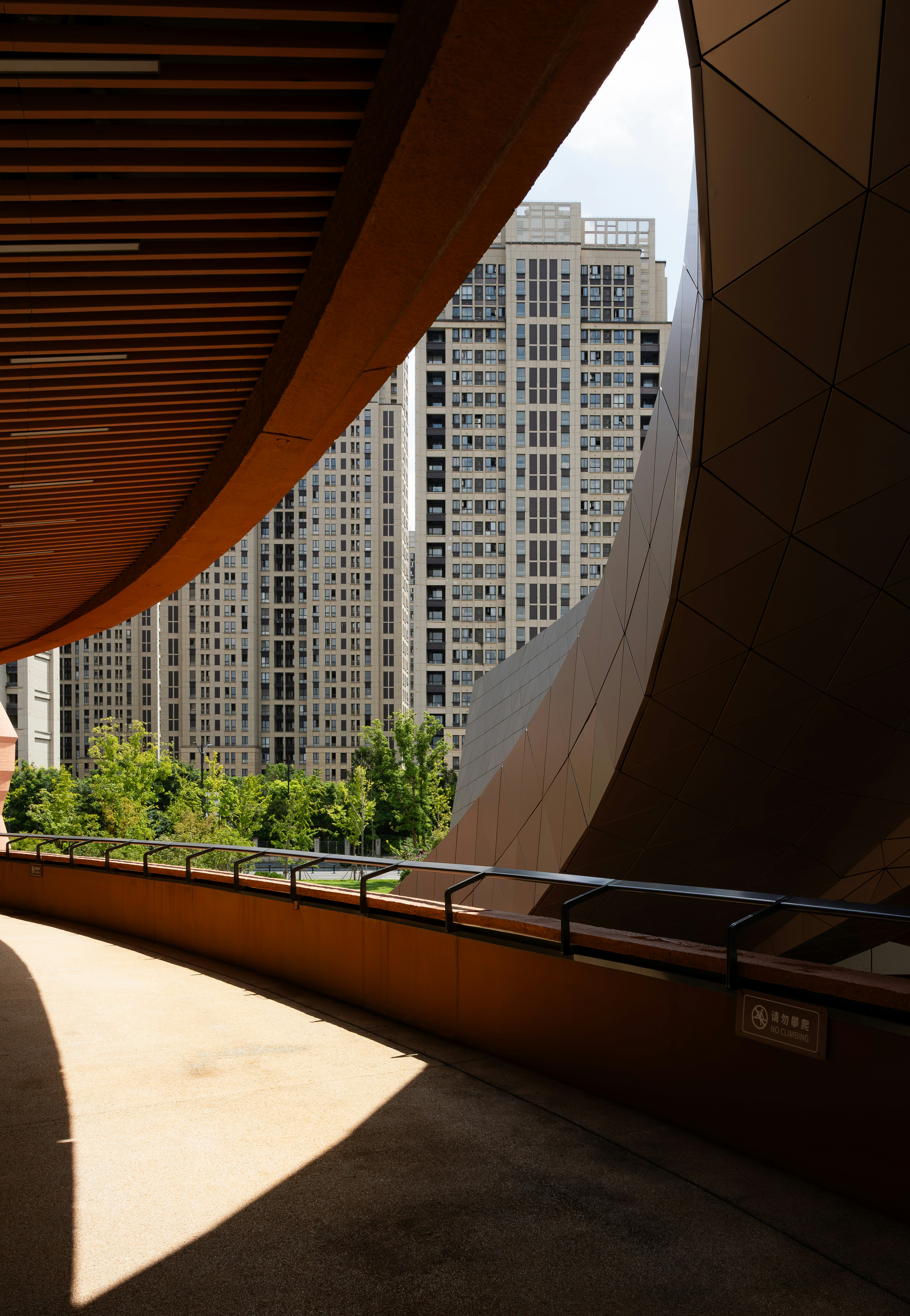 Sunlit urban canyon framed by sweeping metal curves and a distant cluster of high-rise buildings, with a pedestrian pathway in shadow. Modern architectural elements contrast with greenery and bright sky.