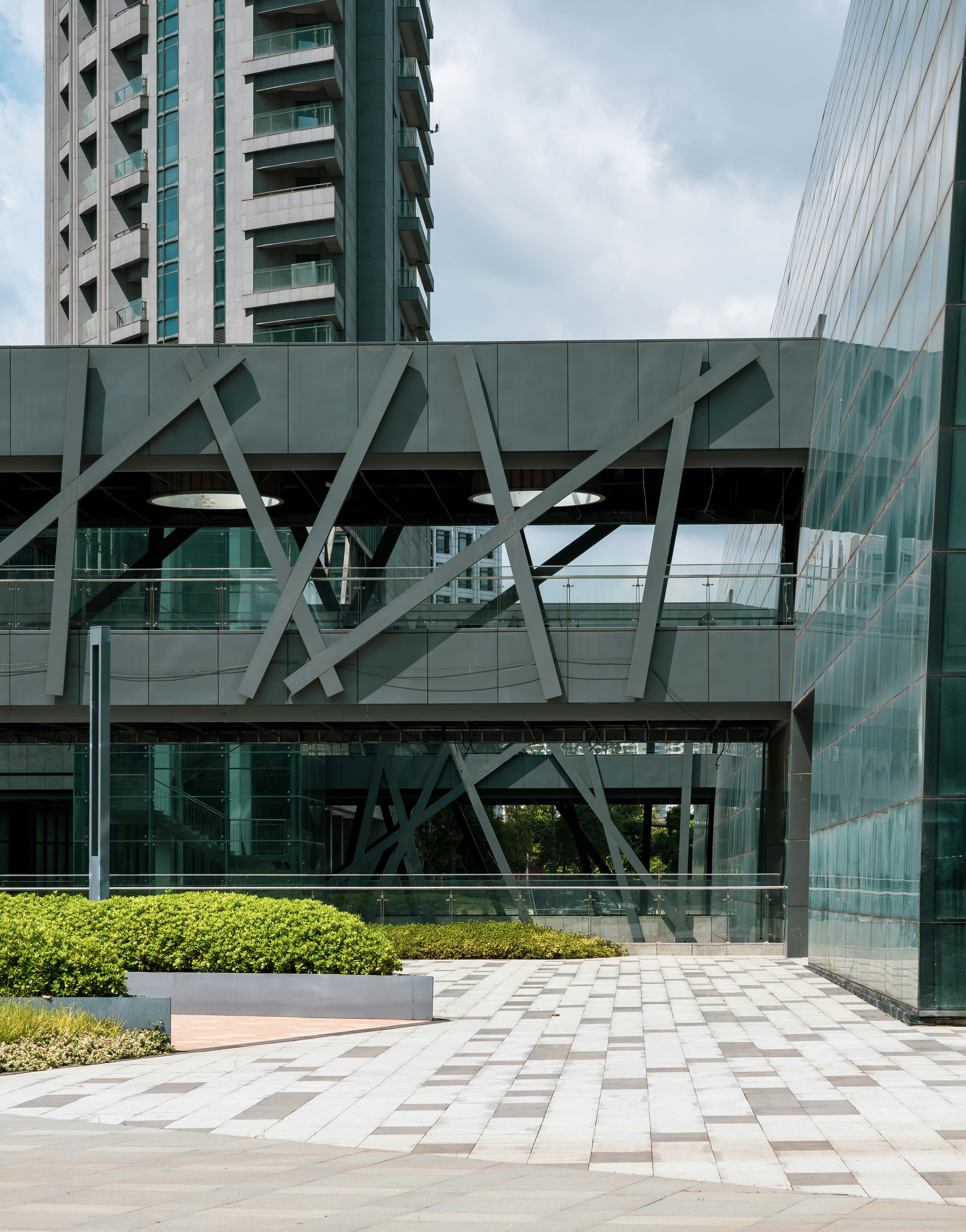 Clean architectural detail of a modern glass and steel structure with crisscross supports and a tiled plaza in the foreground.