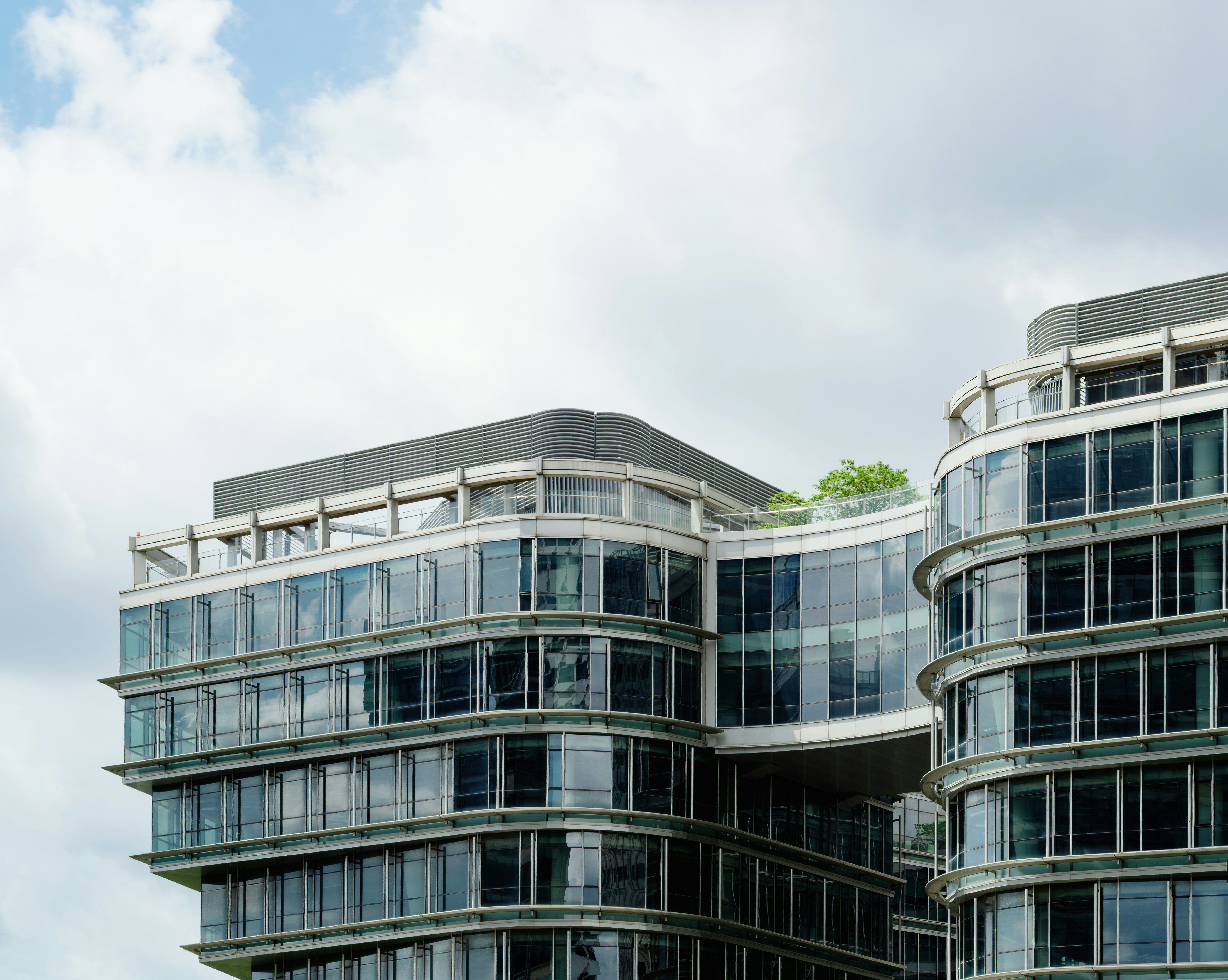 Sleek glass-clad corner of a contemporary office building with rounded balconies and reflective panels, emphasizing architectural geometry.