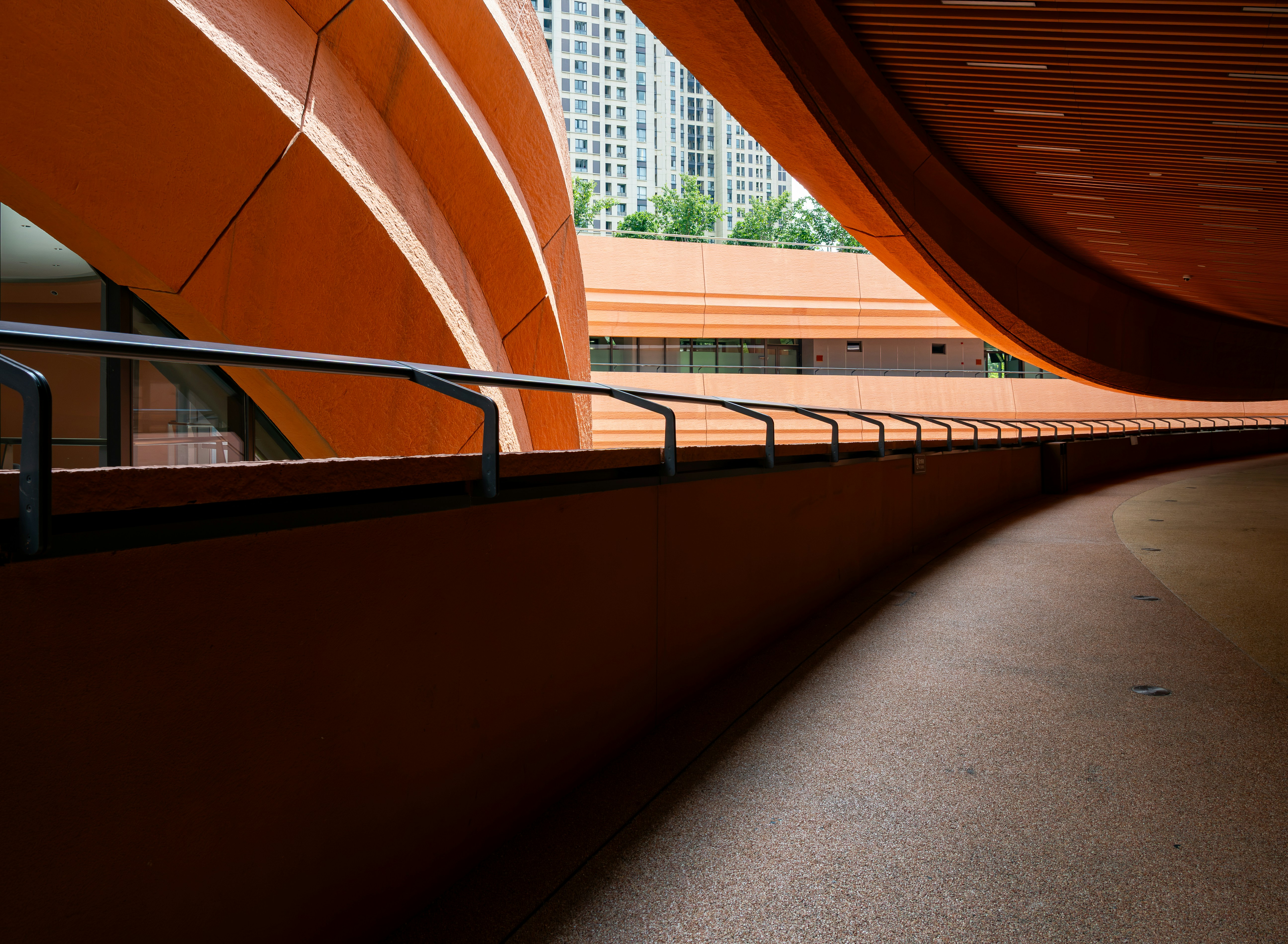 Sleek orange architectural curve with a walkway and railing leading toward a bright opening; minimalist composition emphasizing forms and lines. This is a photograph of an architectural environment.