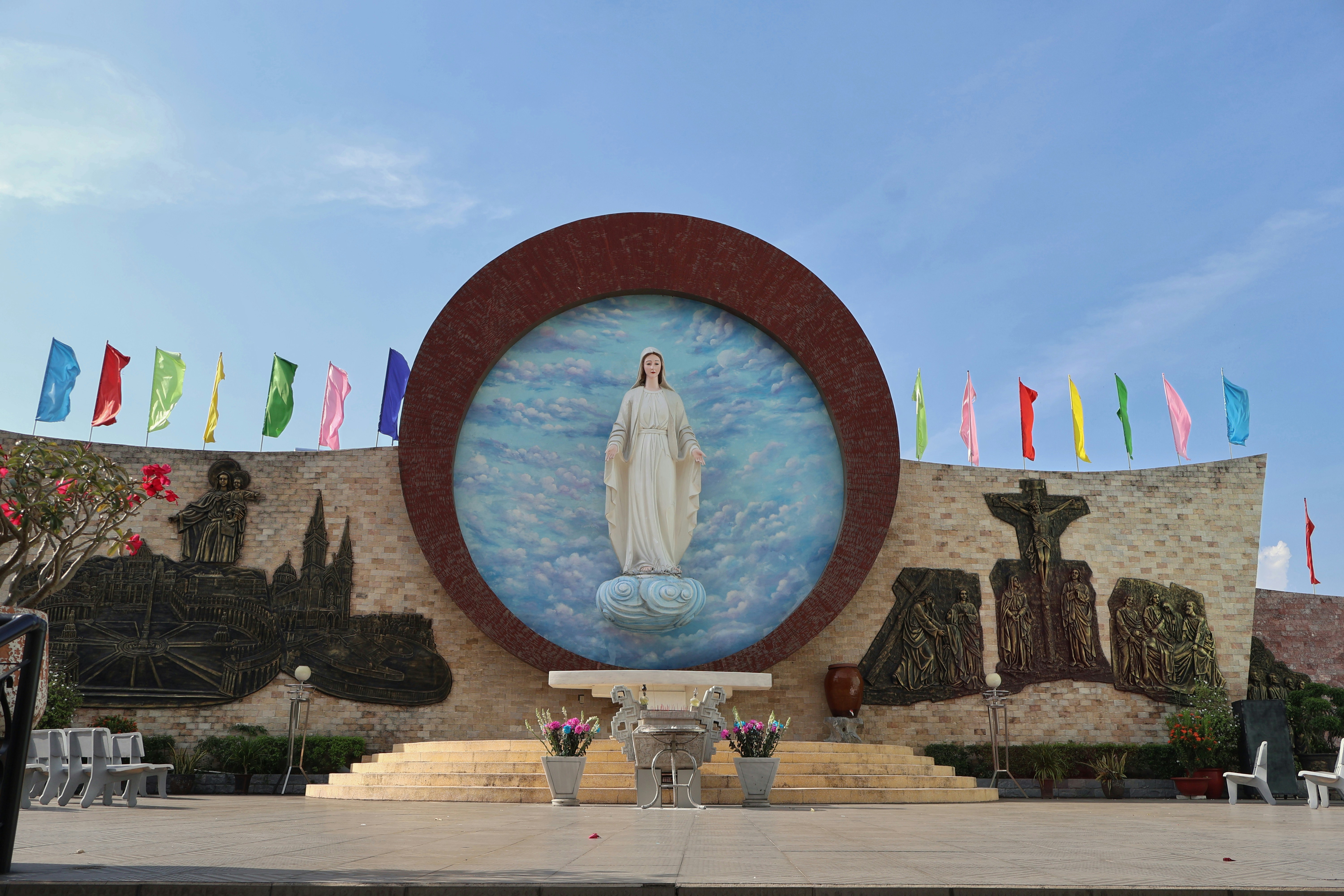 Circular niche with a luminous statue of a serene figure centered against a blue sky, flanked by sculpted panels and colorful flags on a stone plaza.