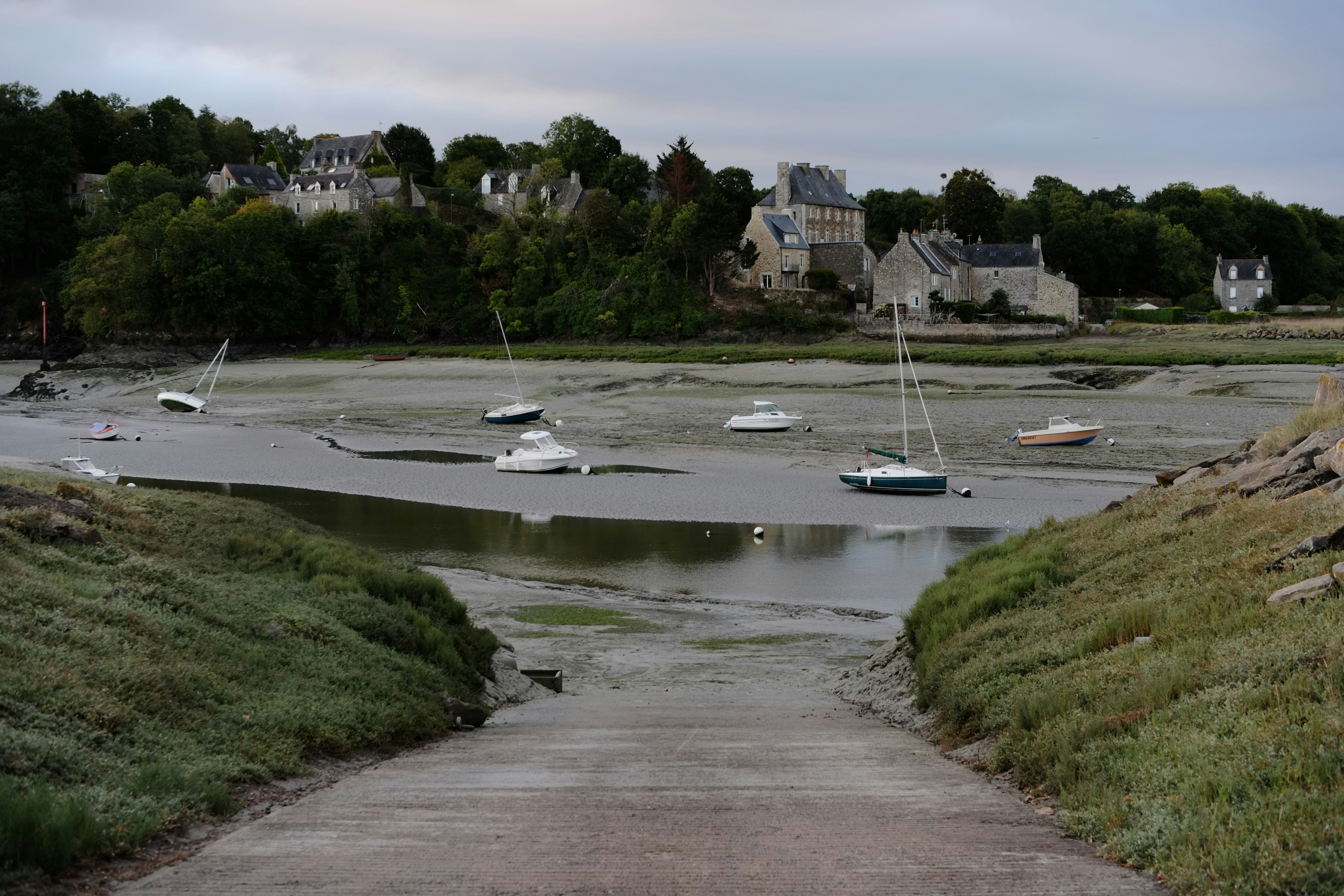 Sailboats anchor in a shallow tidal inlet with a village in the background; the scene emphasizes calm waters and subdued colors in a coastal setting.