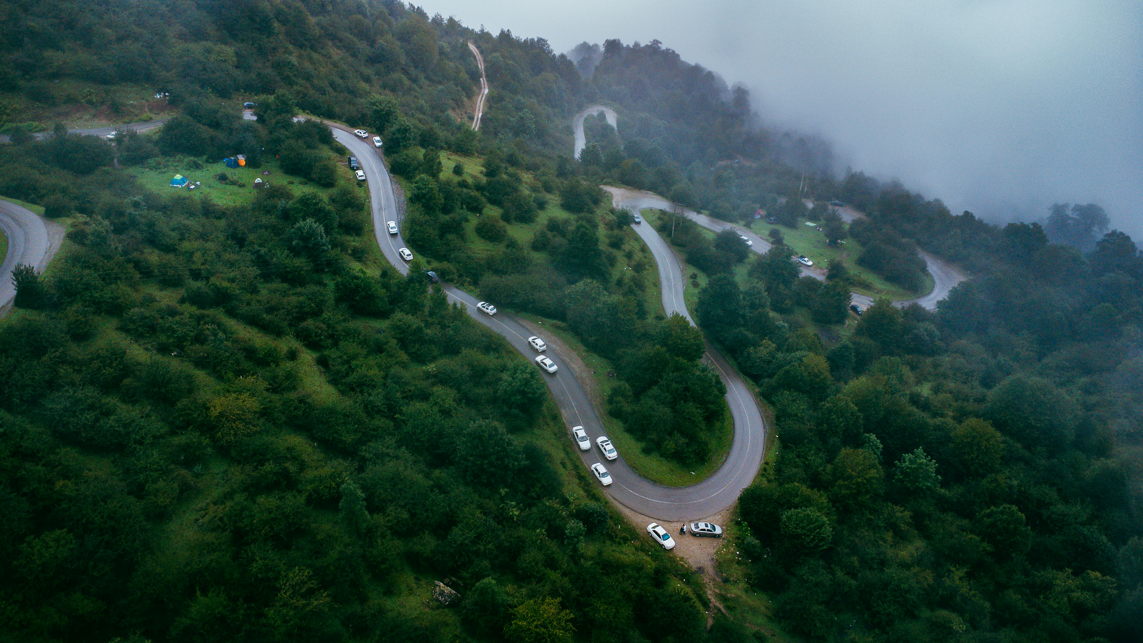 Aerial view of a winding mountain road cutting through dense forest, with misty clouds hugging the tree line and a line of vehicles following the bend.