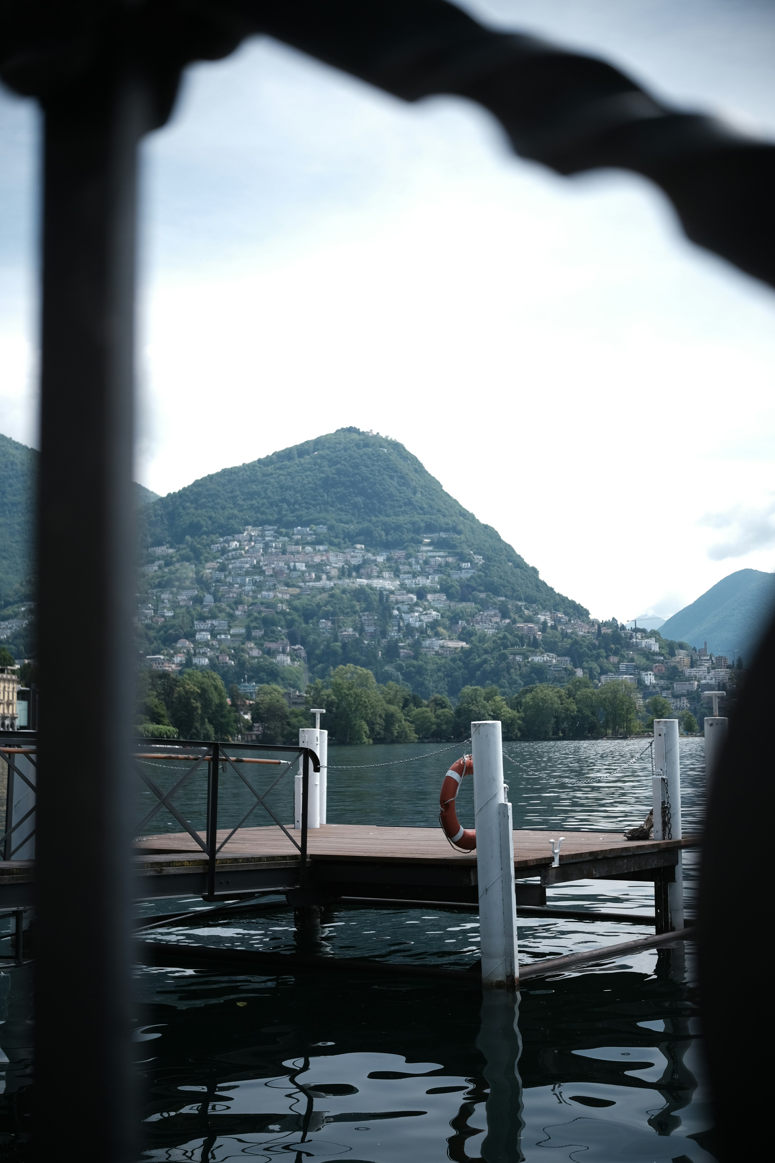 A tranquil lakeside scene viewed through a boat dock, with a hillside village on a lush mountain slope reflected in calm water. The image captures a quiet moment by the pier, featuring a lifebuoy and moored posts.