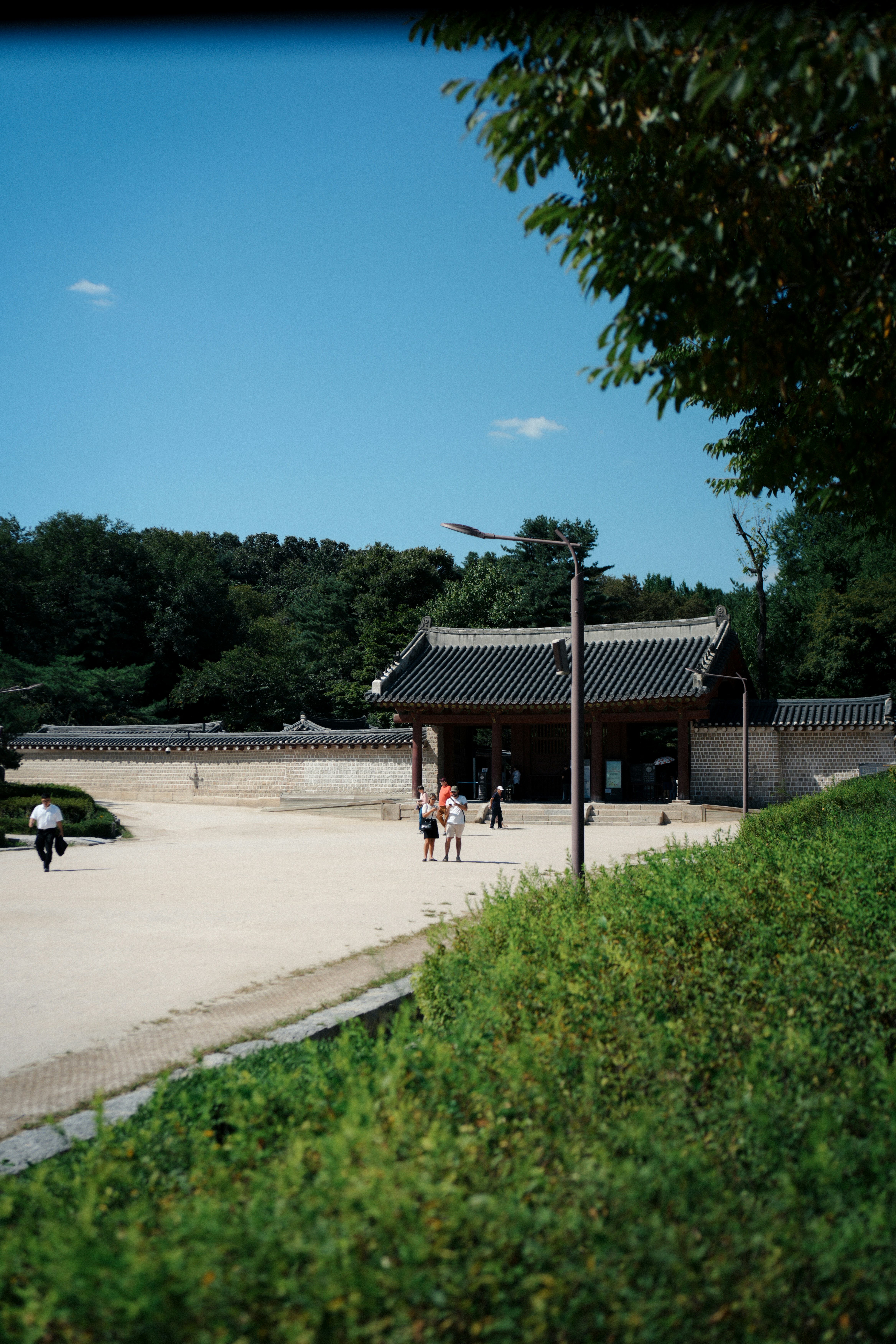 Visitors stand at the entrance of a traditional tiled pavilion flanked by a low wall and lush greenery. A wide plaza leads to the structure with a clear blue sky above.