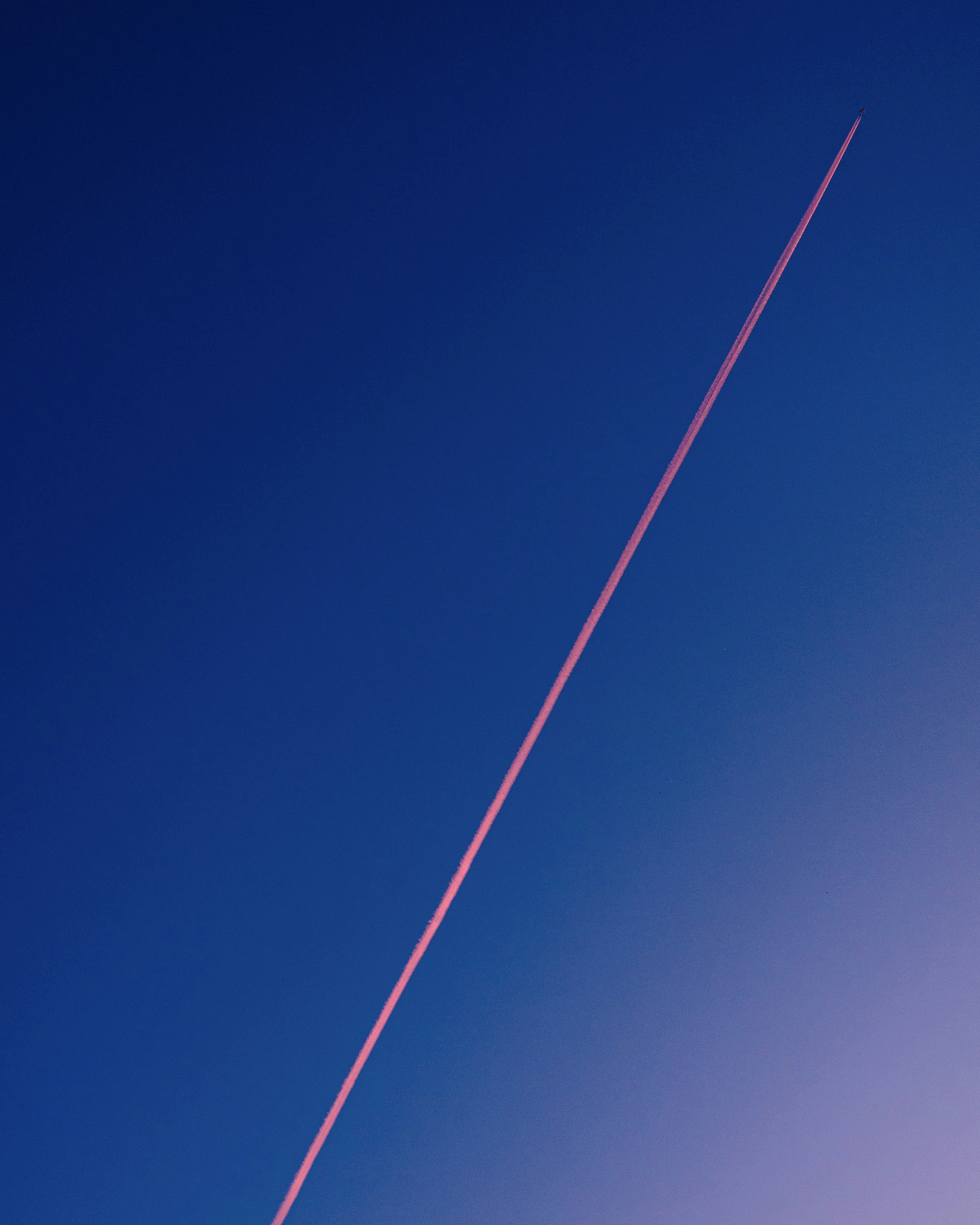 A vivid pink contrail cuts diagonally across a deep blue sky, fading toward a hazy gradient at the lower right; the composition emphasizes the line against a minimalist backdrop.