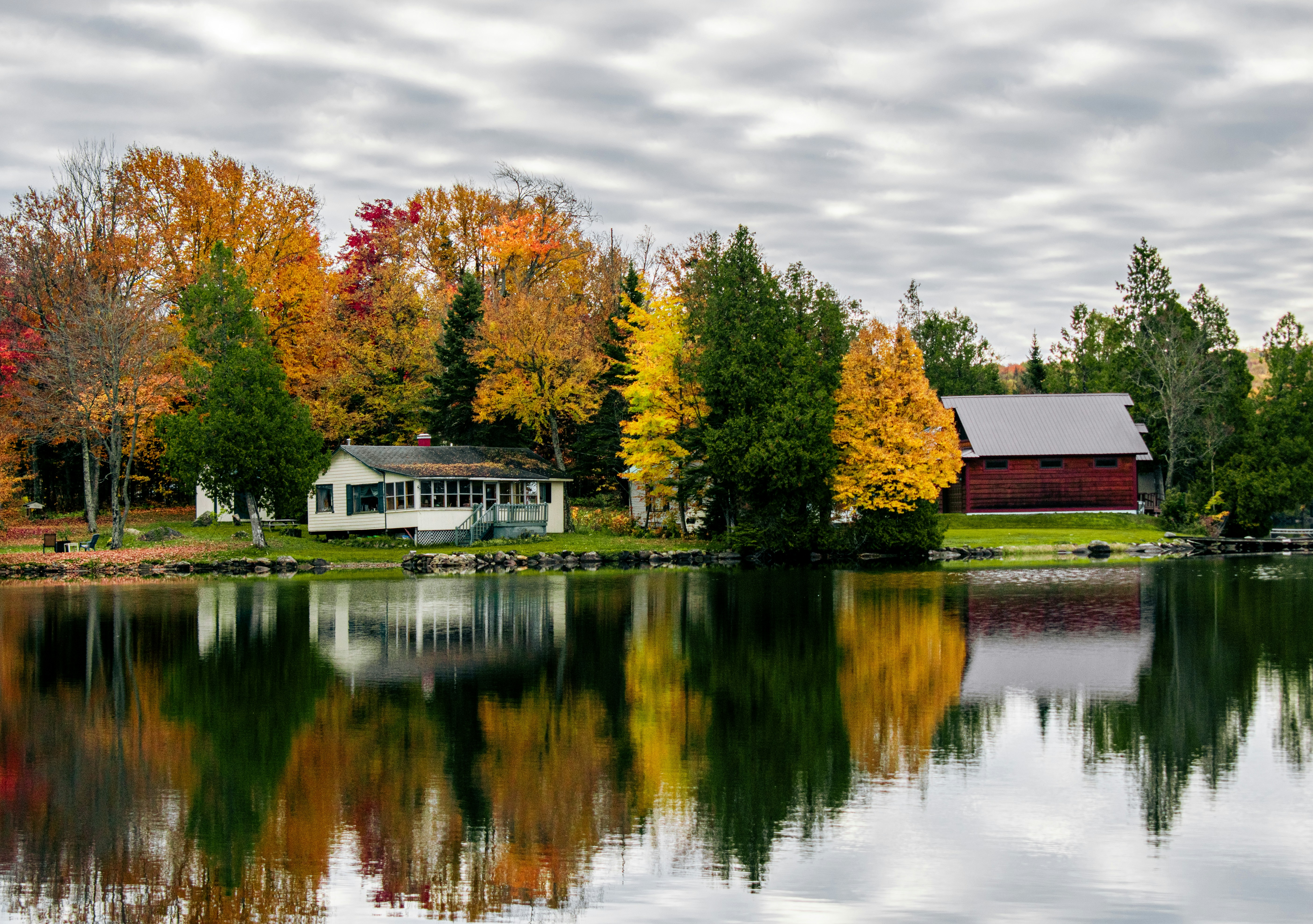 Pastel autumn trees line the tranquil lakeshore with a white cottage and red barn reflected in glassy water.